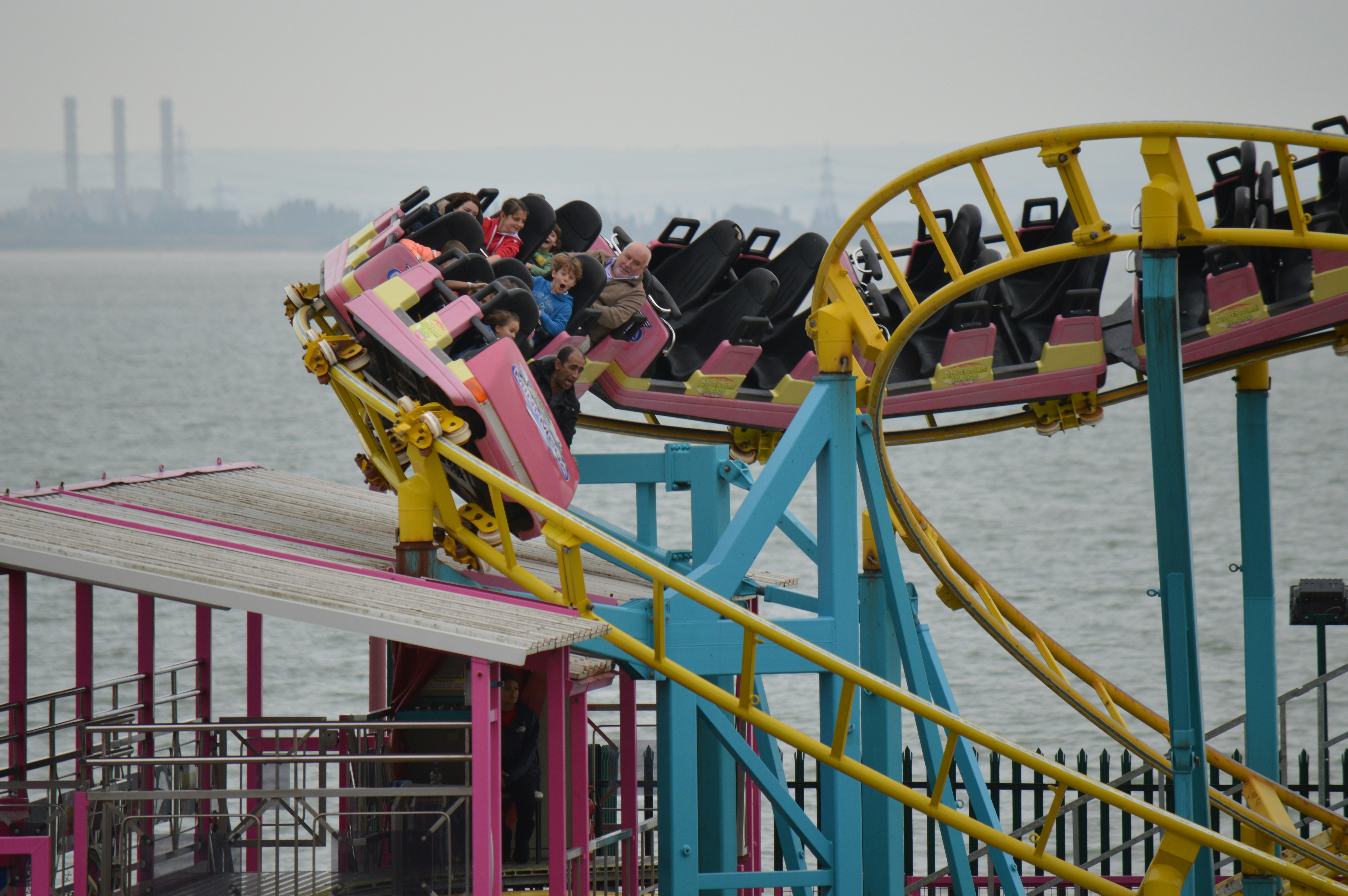 Colorful roller coaster with excited riders leaning forward as it approaches a steep drop, overlooking a calm sea.