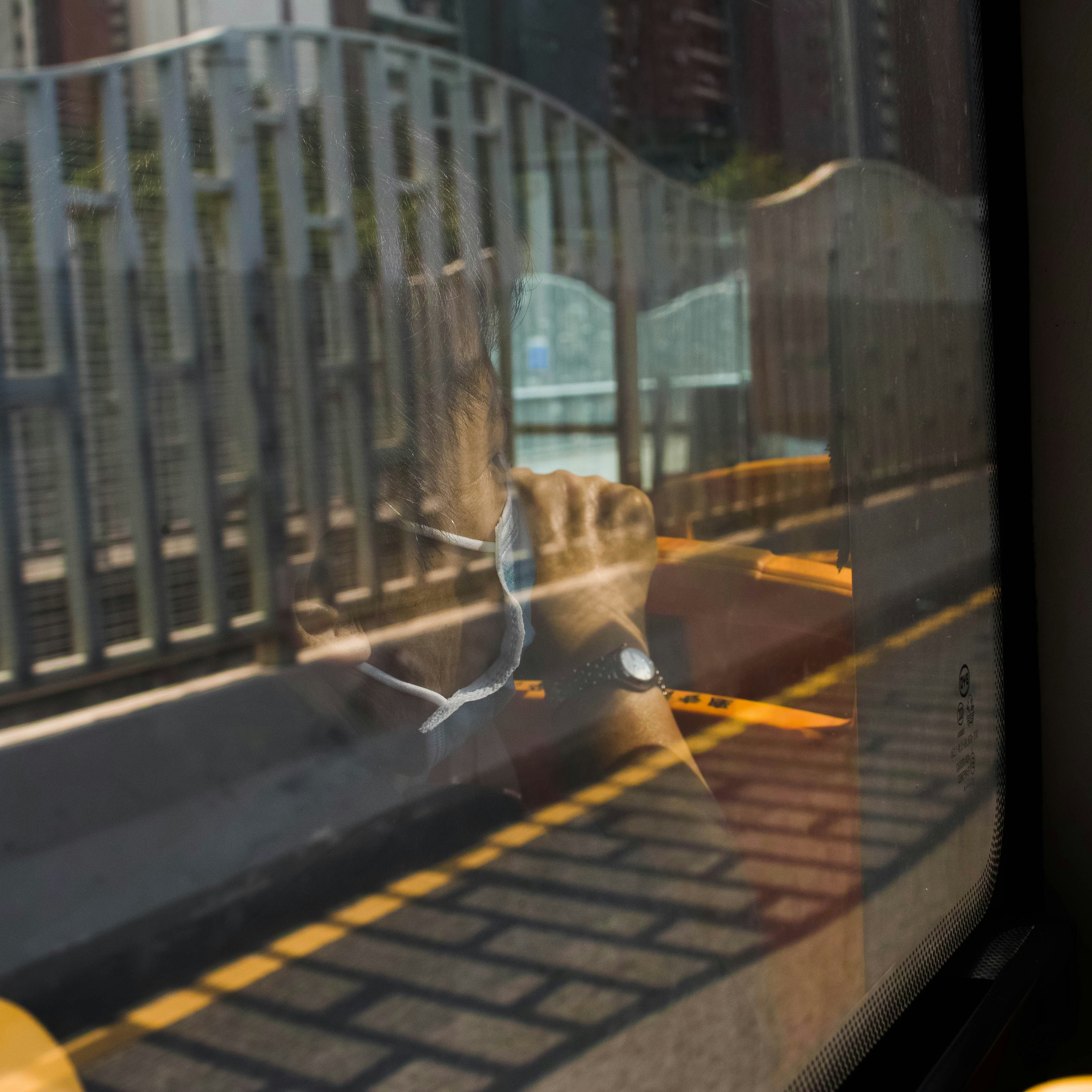 Reflection of a masked person seen in a bus window, with metal railing and yellow road markings visible outside. This photograph captures a quiet urban moment through glass.