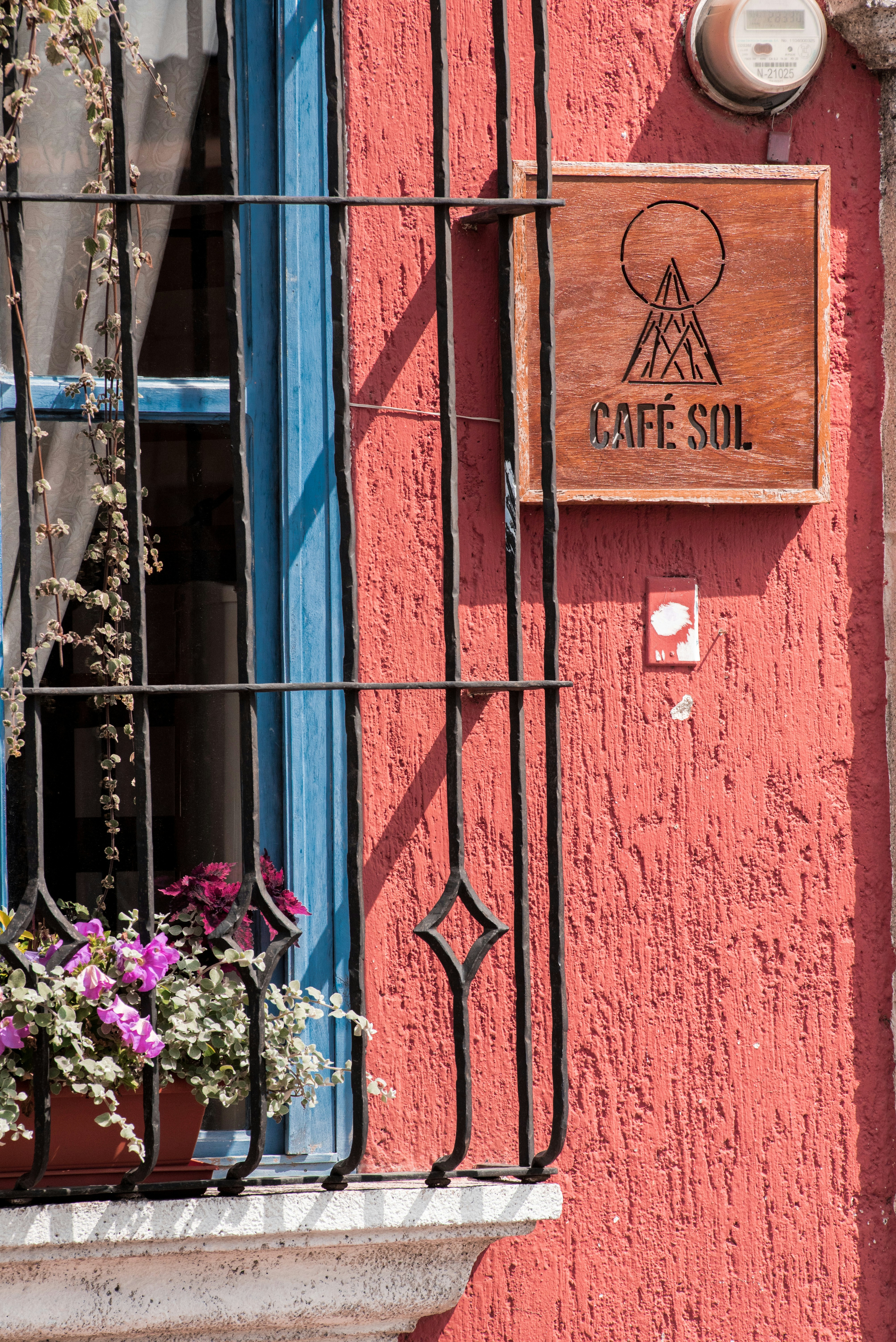 A wooden sign reading 'CAFÉ SOL' hangs beside a vibrant blue window adorned with blooming flowers, set against a textured red wall.
