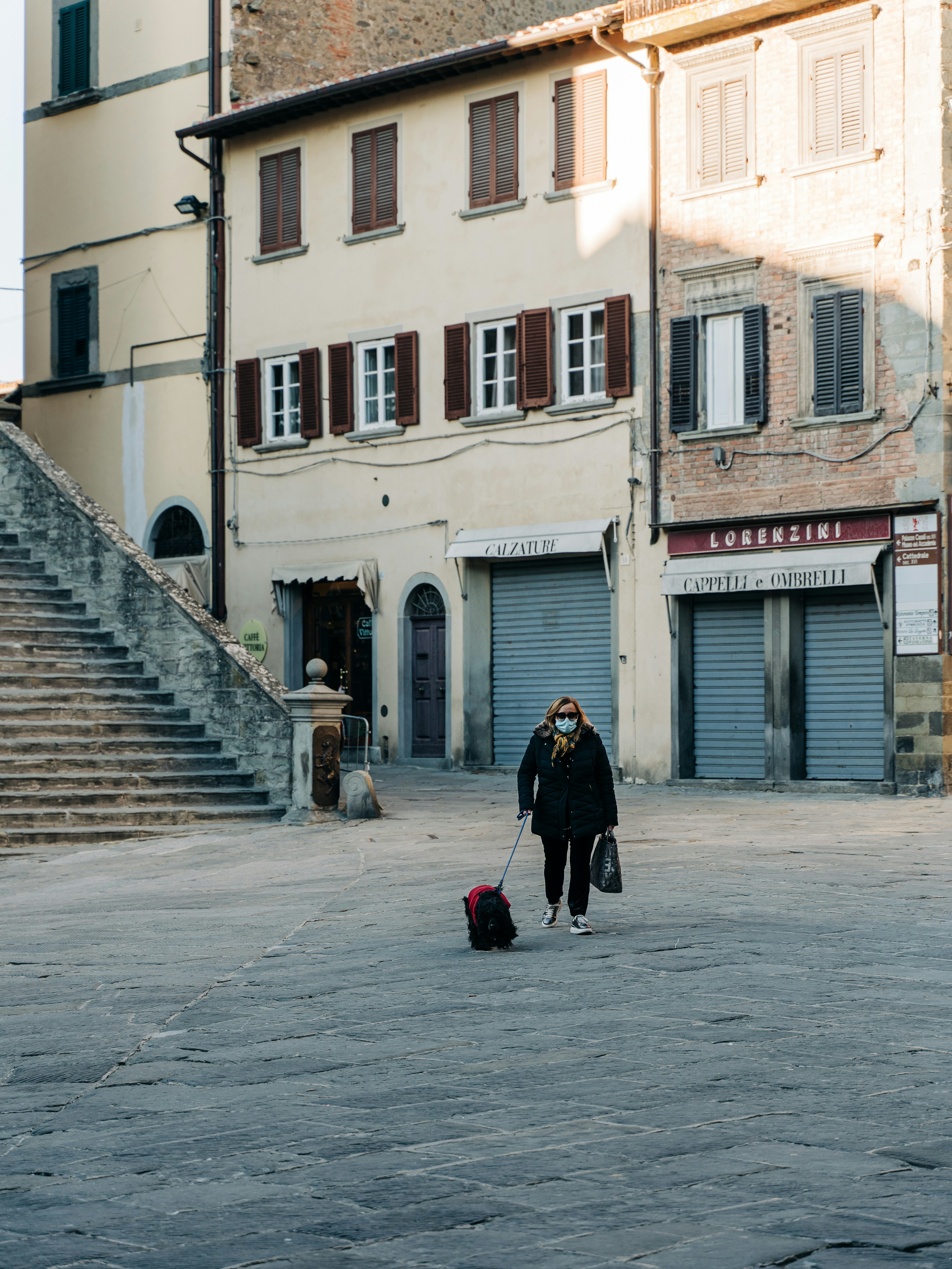 Woman walking a small dog in a quaint Italian street, flanked by historic buildings and closed storefronts. Morning light casts soft shadows on the cobblestone pavement.