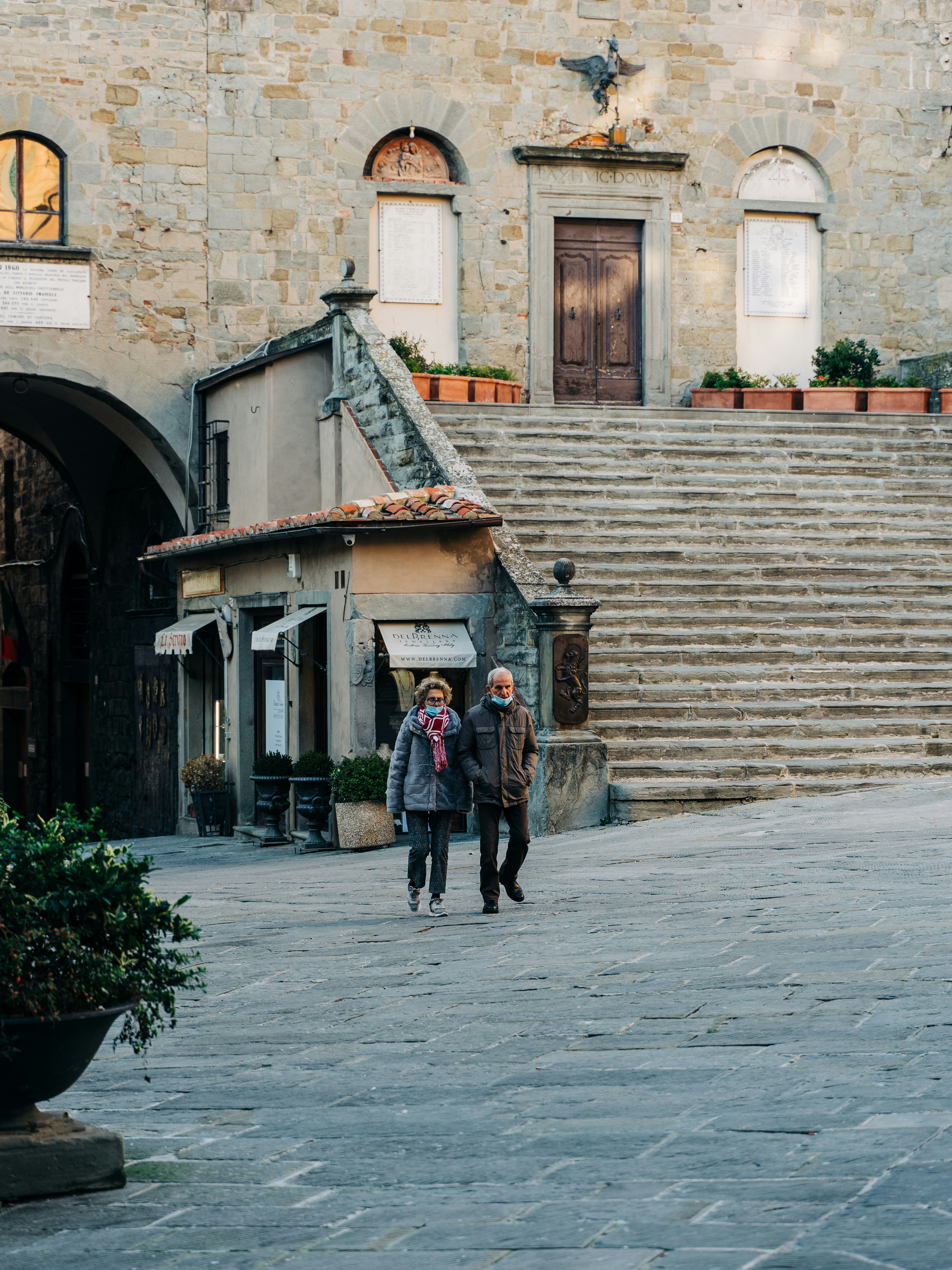 Two masked wanderers descend a sunlit stone stair at a historic town square