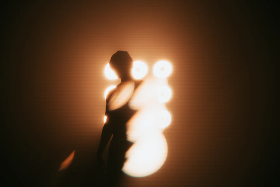 A silhouetted person stands in front of a vertical row of bright warm lights, with strong blur and haloing around the highlights. The scene feels moody and abstract, with the subject centered slightly left.