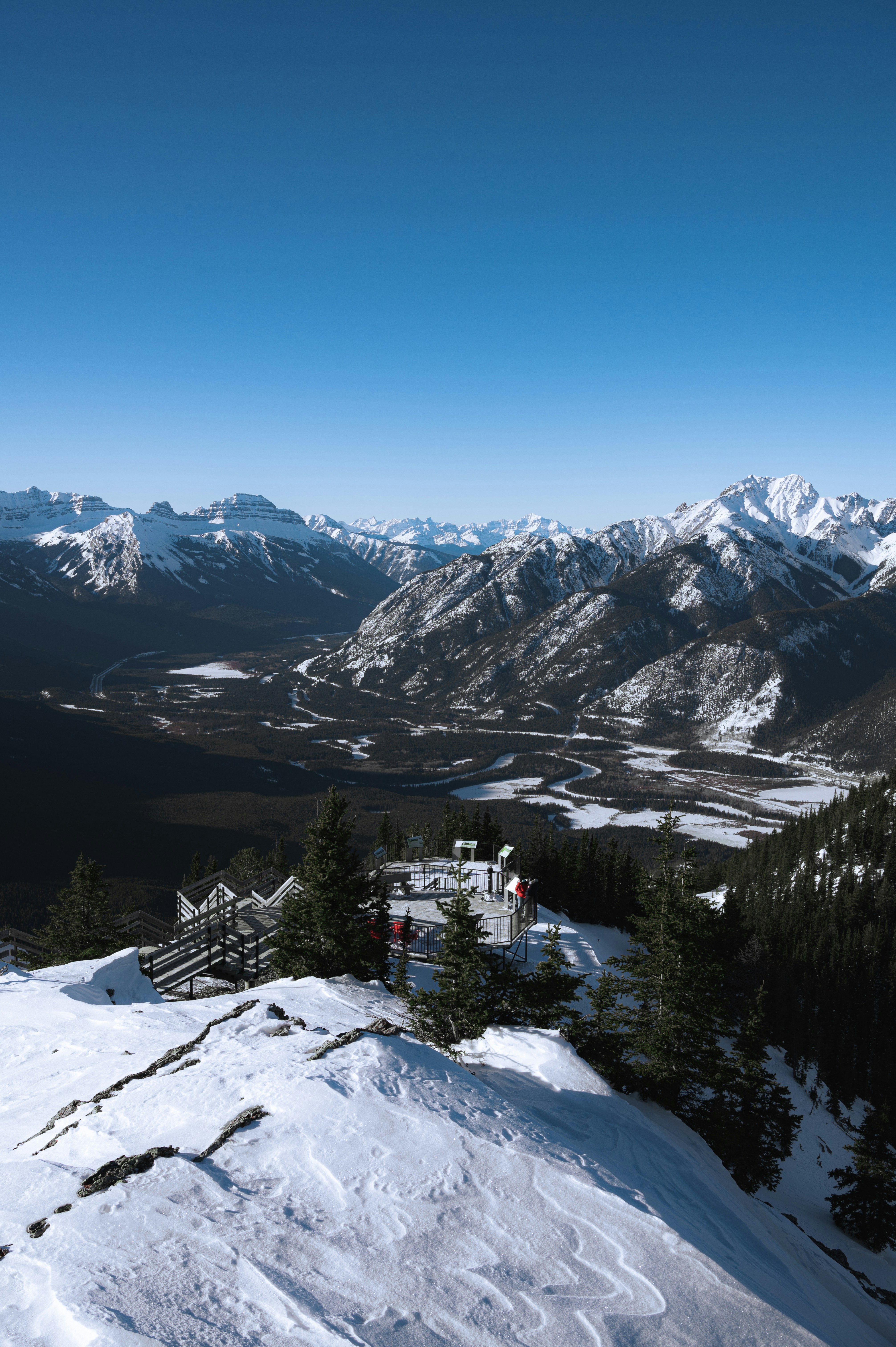 Snow-covered mountain peak overlooking a winding river valley, with a viewing platform nestled among evergreen trees. A clear blue sky enhances the tranquil winter landscape.