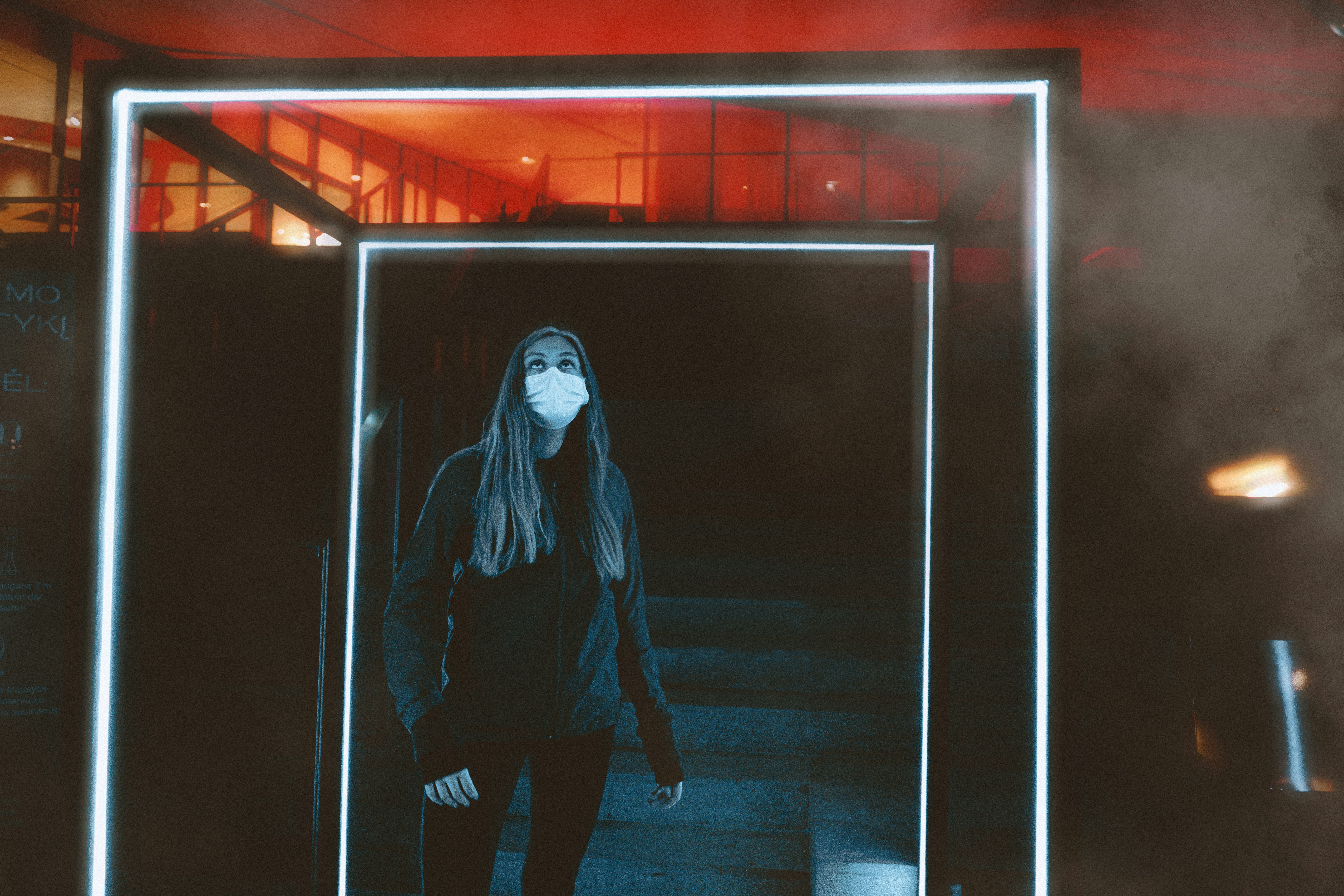A young woman in a mask stands within a glowing rectangular frame, surrounded by a moody ambiance. The contrasting lights create a striking visual narrative.