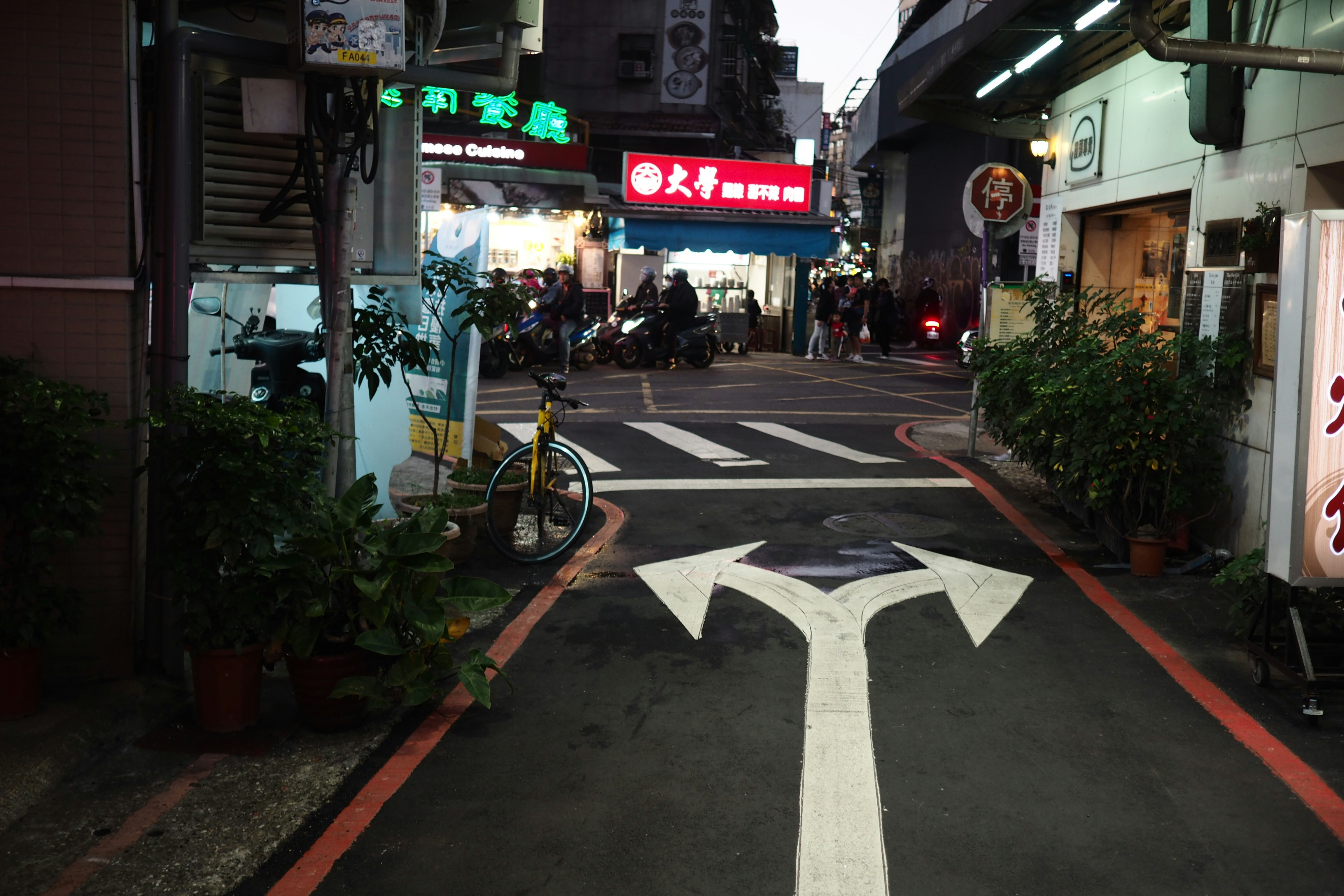 A narrow urban alleyway featuring a prominent white arrow marking the direction, surrounded by greenery and illuminated storefronts. The scene captures the essence of city life at dusk.