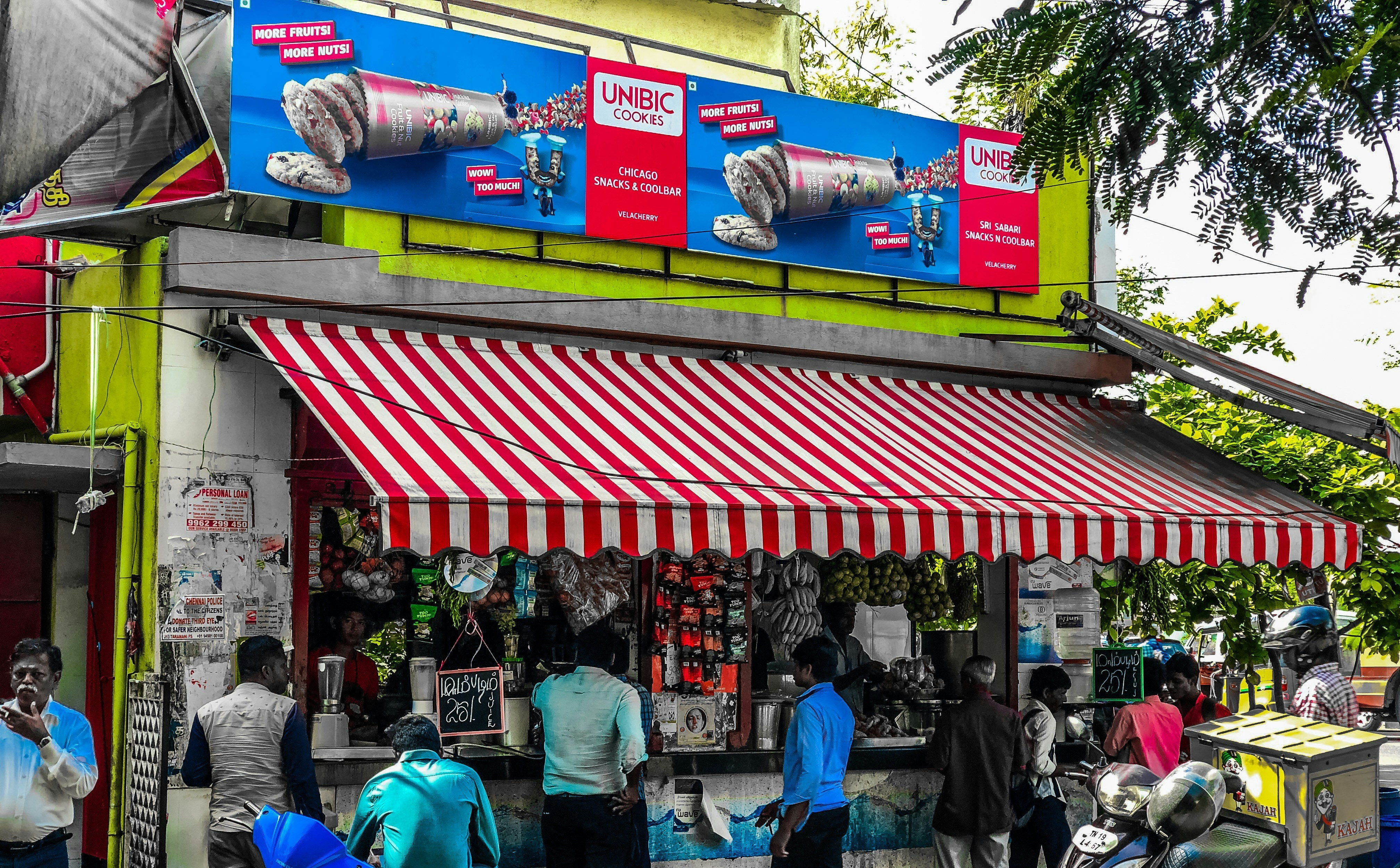 A lively corner store adorned with colorful signage and striped awnings, bustling with customers. The shop showcases a variety of goods and local products.