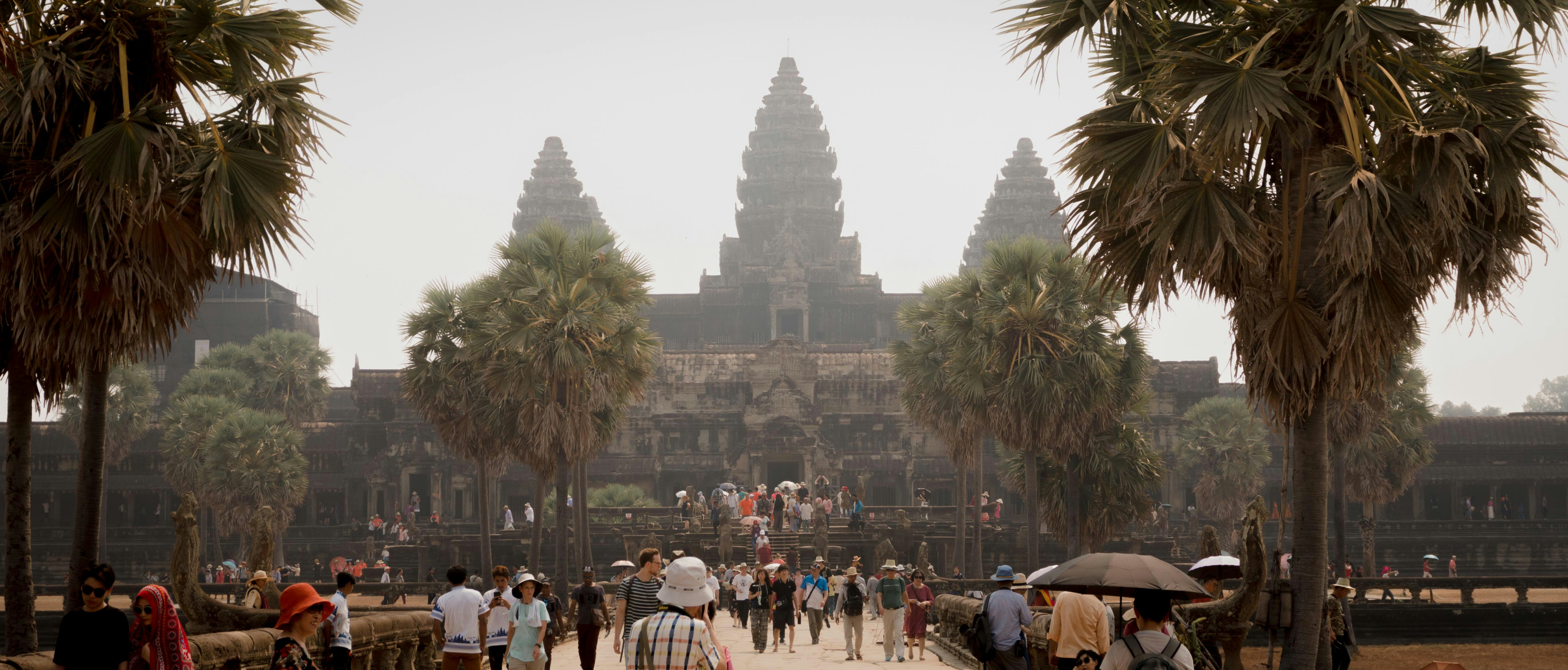 Crowd of visitors exploring the majestic Angkor Wat temple complex, framed by lush palm trees under a hazy sky.
