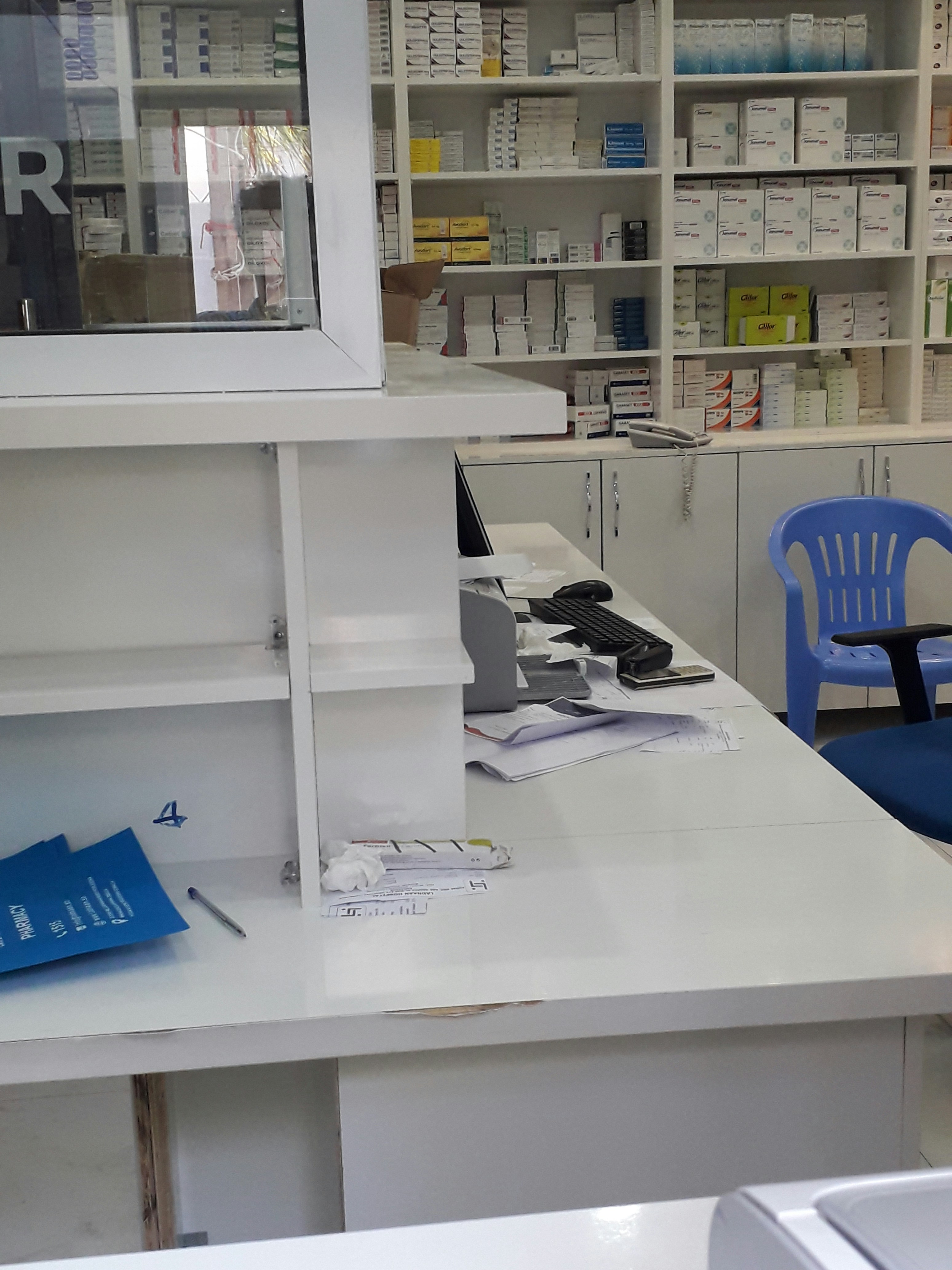 Pharmacy counter with a computer setup, documents, and a blue plastic chair, showcasing the organized environment of a healthcare facility.