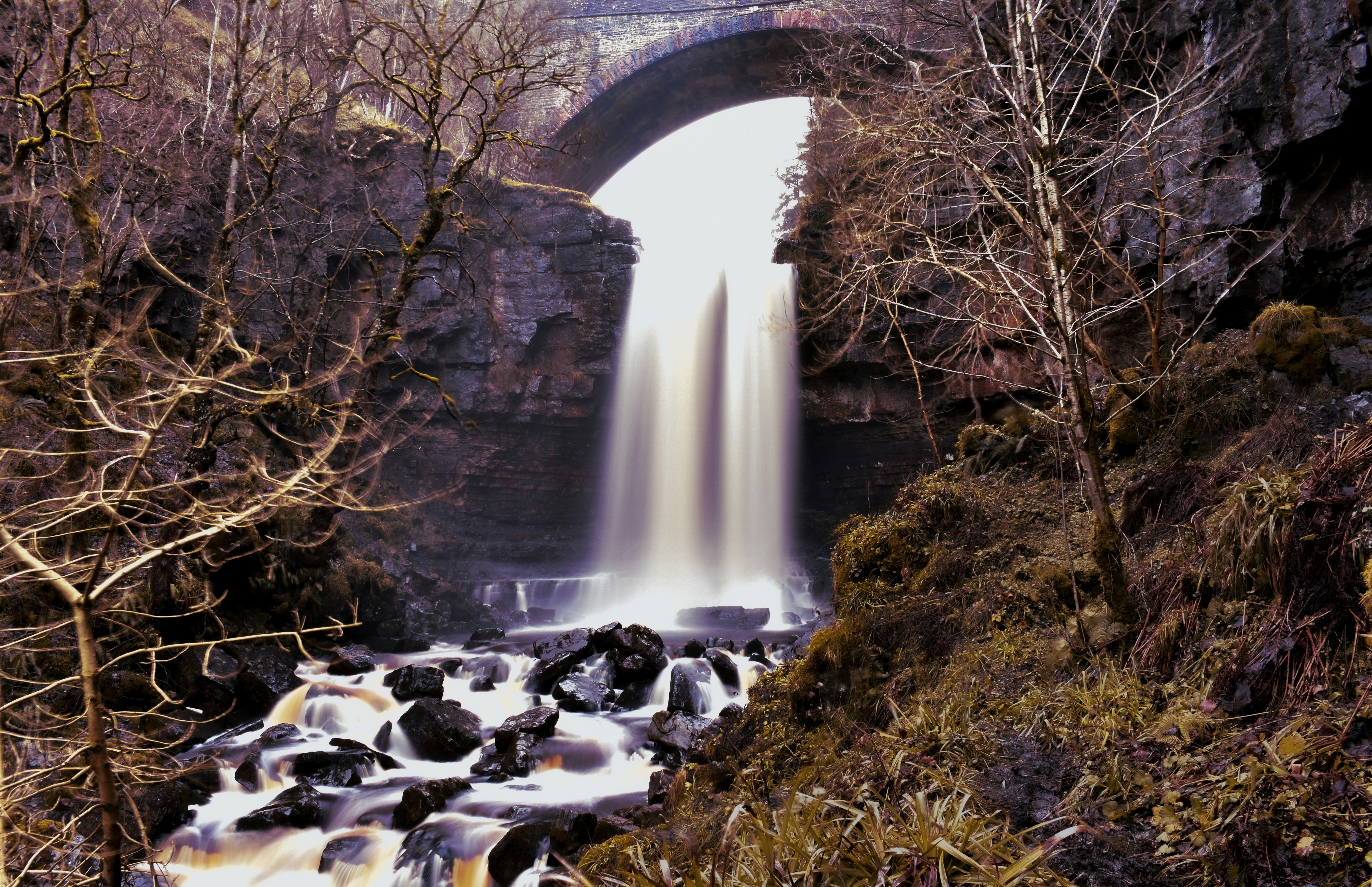 Majestic waterfall cascading under a stone bridge, surrounded by rugged terrain and sparse trees. The scene captures the serene flow of water amidst a tranquil landscape.
