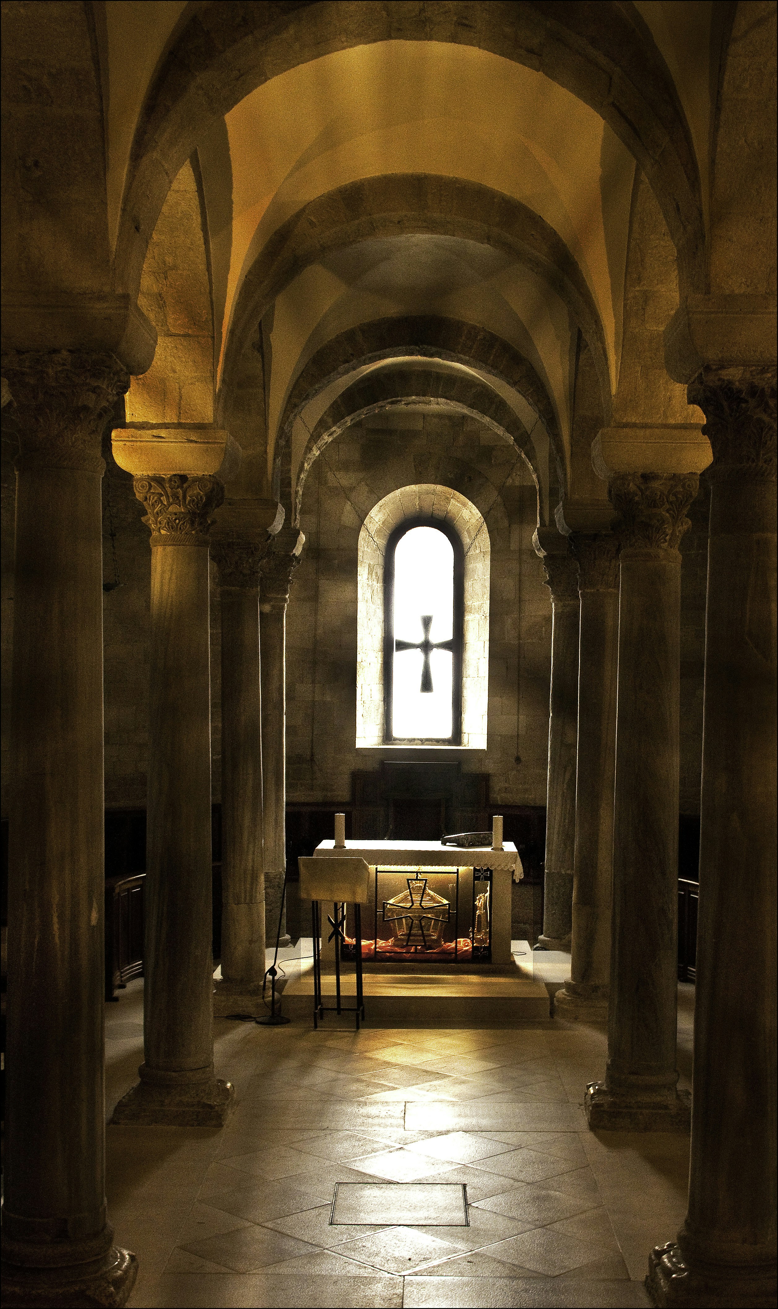 cross in window of crypt altar in trani cathedral | brown wooden bench in a brown concrete building