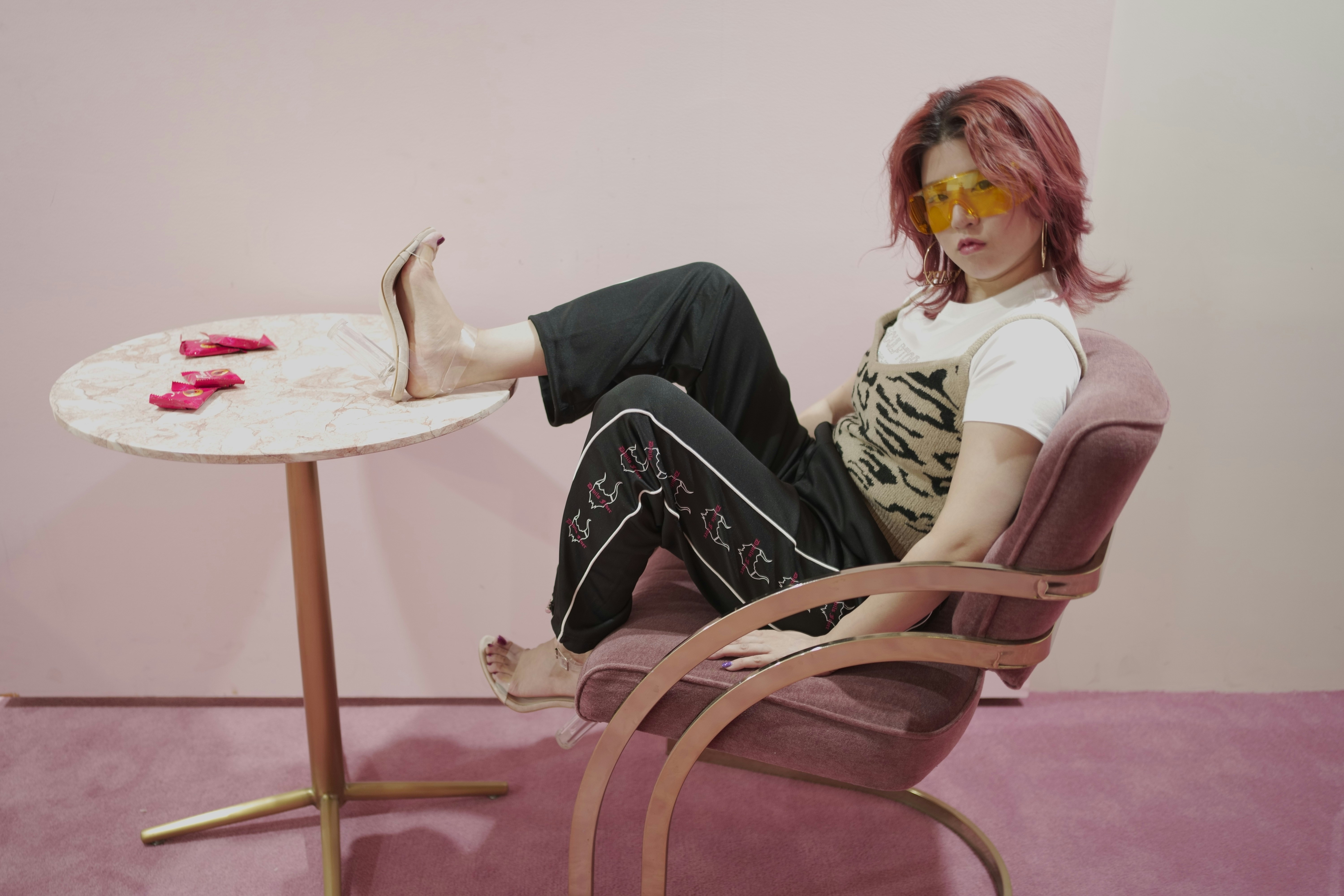 Fashion-forward individual lounging in a stylish chair, showcasing bold attire and vibrant sunglasses against a pastel backdrop.