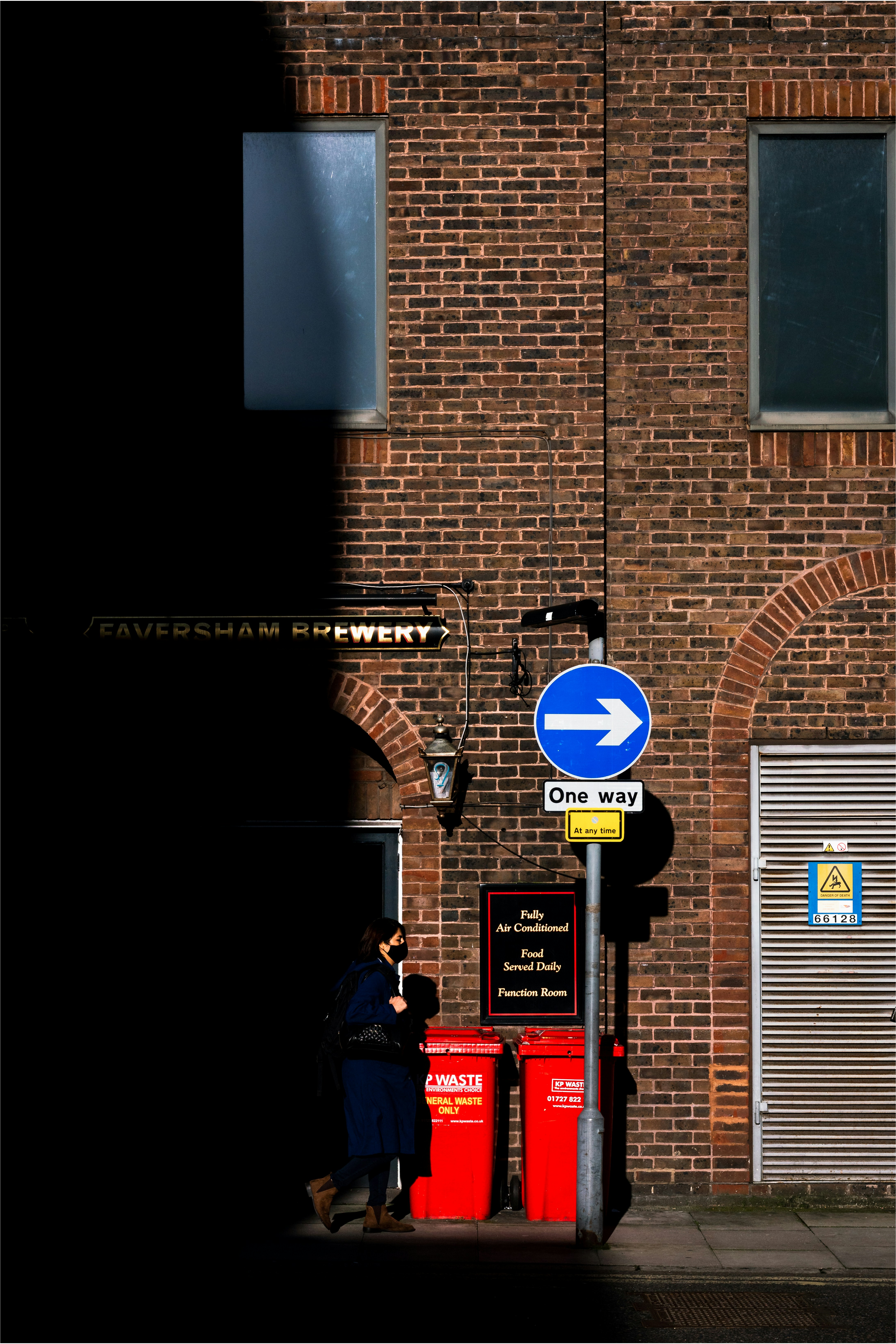 Photograph of a sunlit urban corner featuring a brick building, a blue one-way sign, and a couple near red waste bins. A deep shadow frames the left side, highlighting contrast between light and architecture.