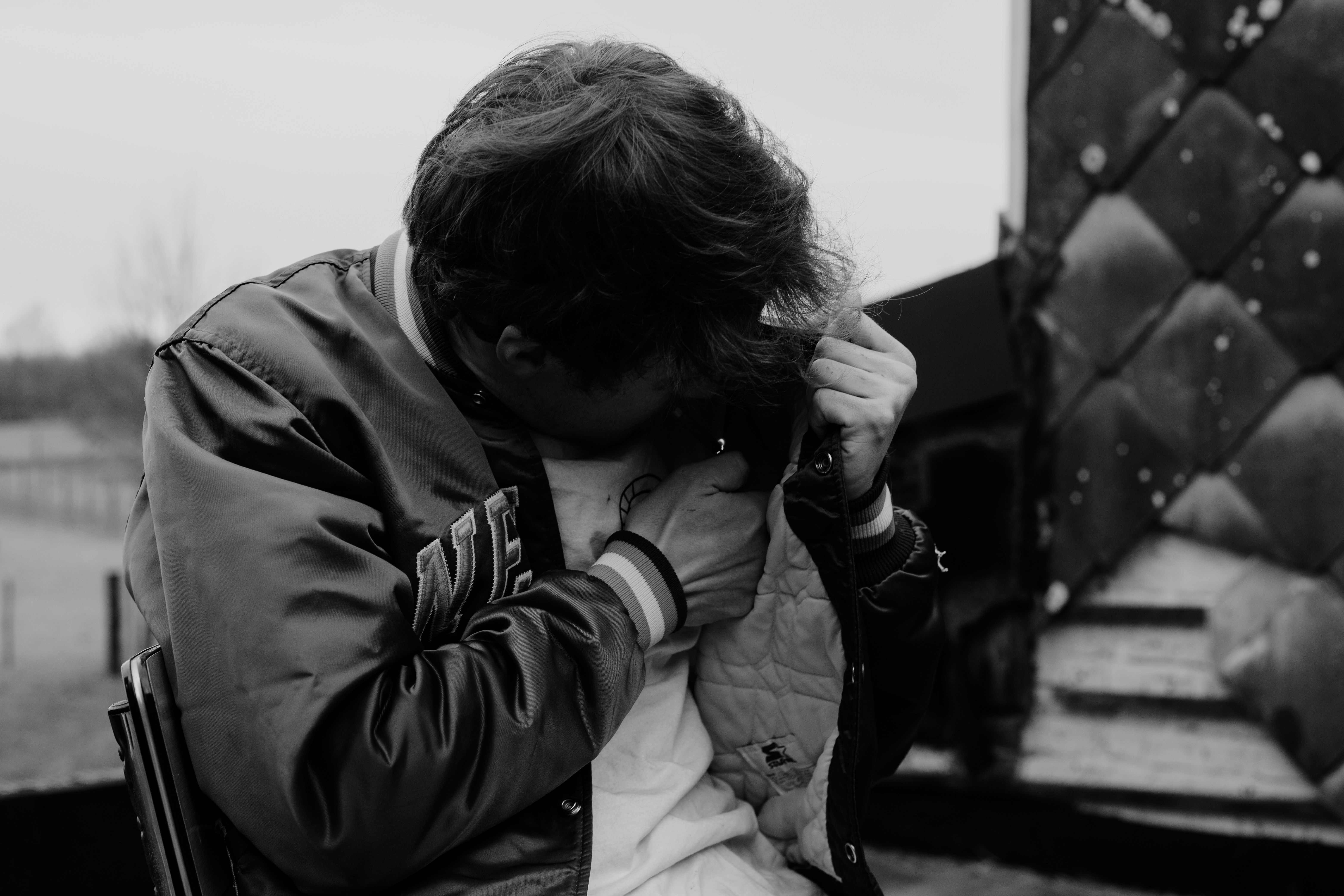 Individual adjusting their jacket in a monochrome setting, surrounded by a rustic backdrop.