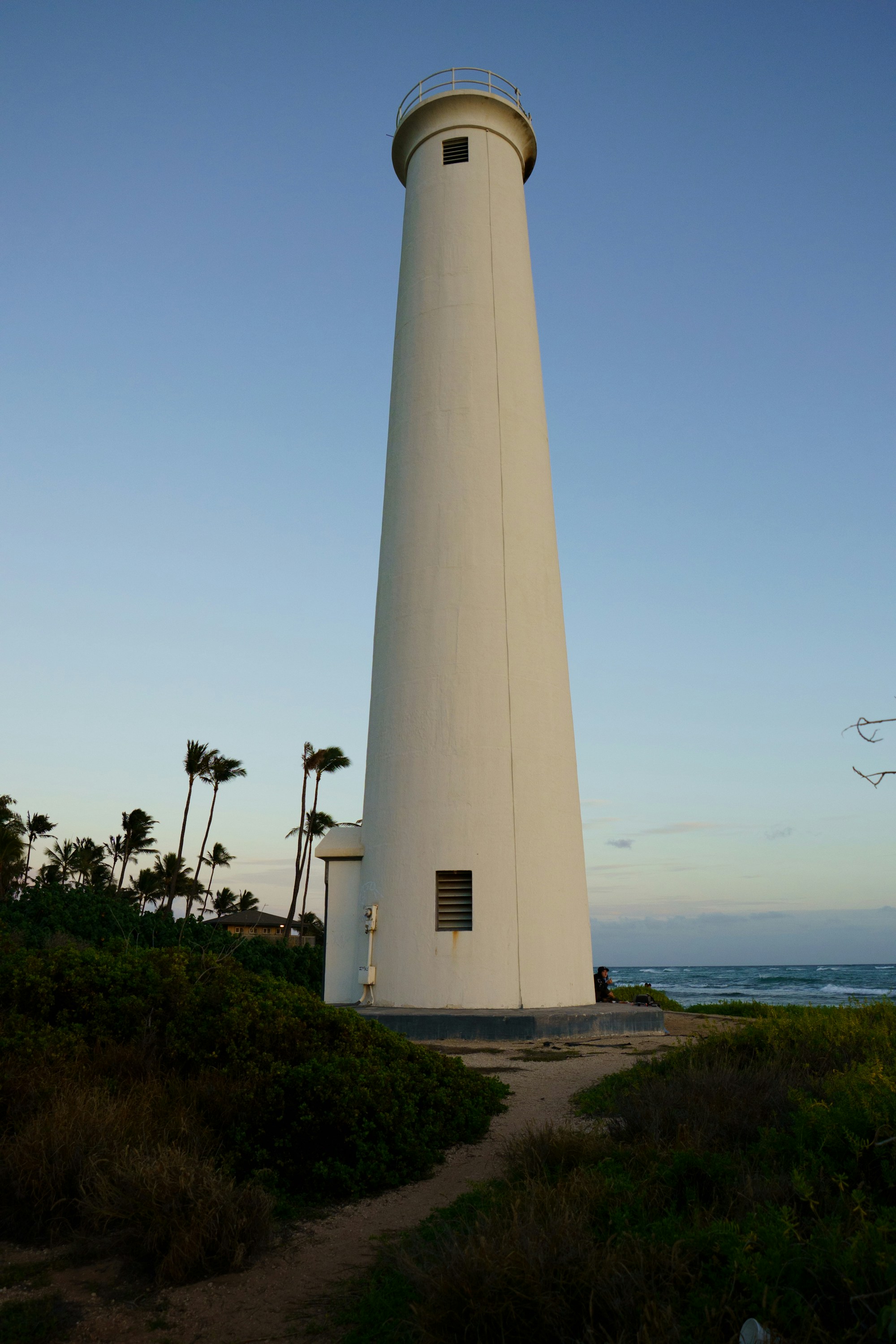 A tall white lighthouse stands proudly on the shore, surrounded by lush greenery and palm trees, with gentle ocean waves in the background.