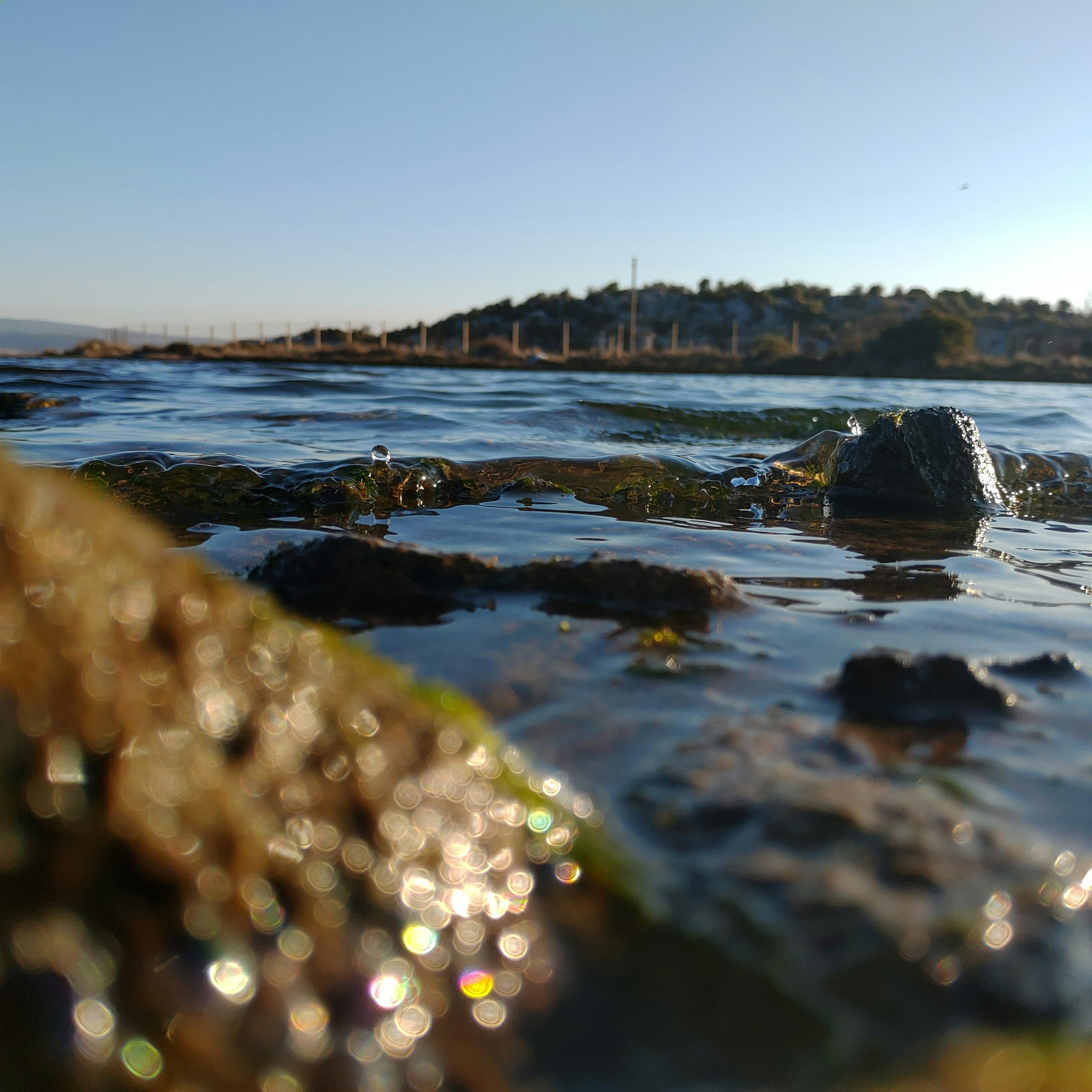 Close-up view of water lapping against rocks, with glimmers of sunlight reflecting off the surface. The scene captures the serene interaction between land and sea.