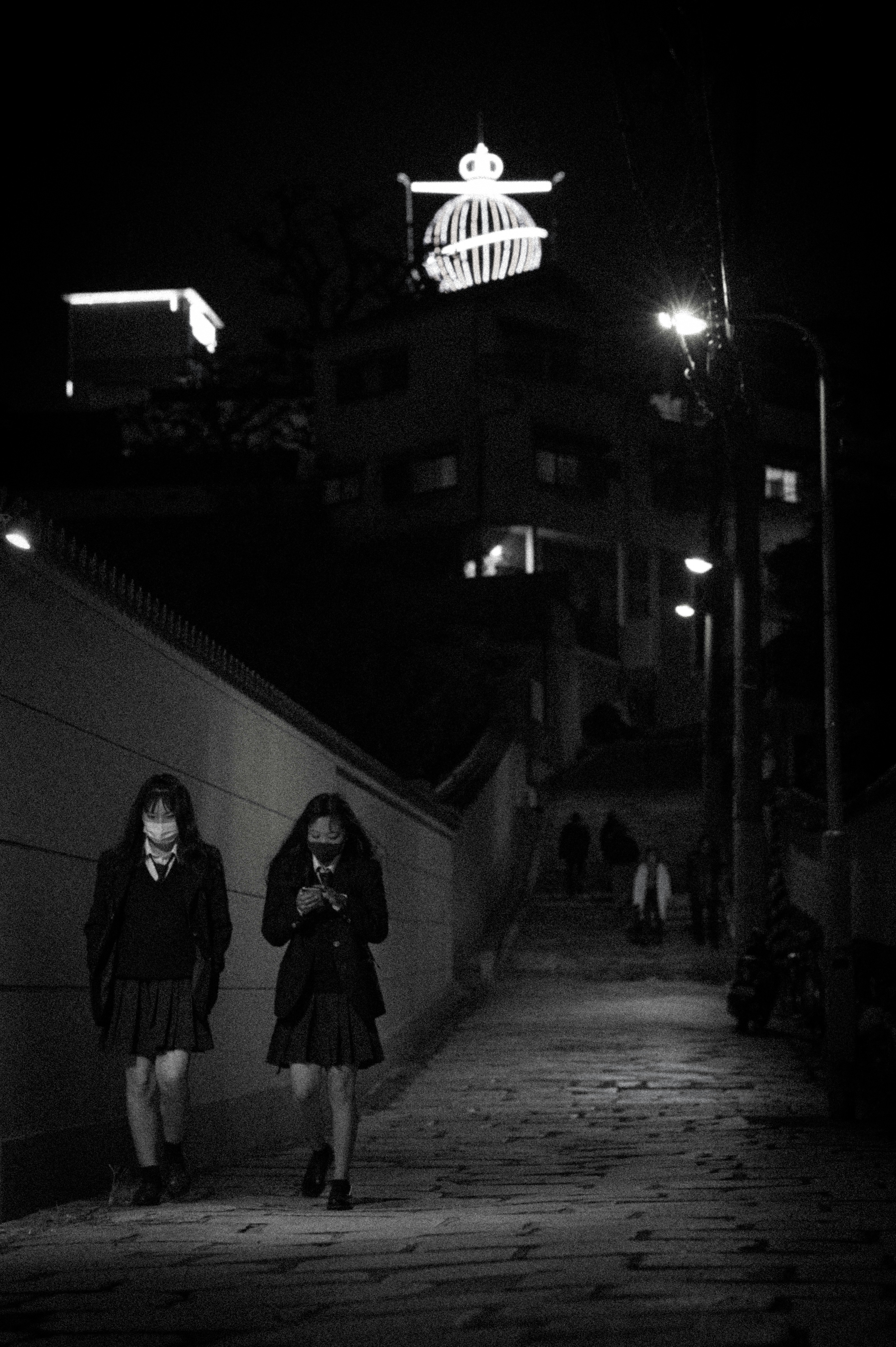 Two students in uniforms walk down a dimly lit street, absorbed in their own worlds. A distinctive illuminated dome looms in the background.