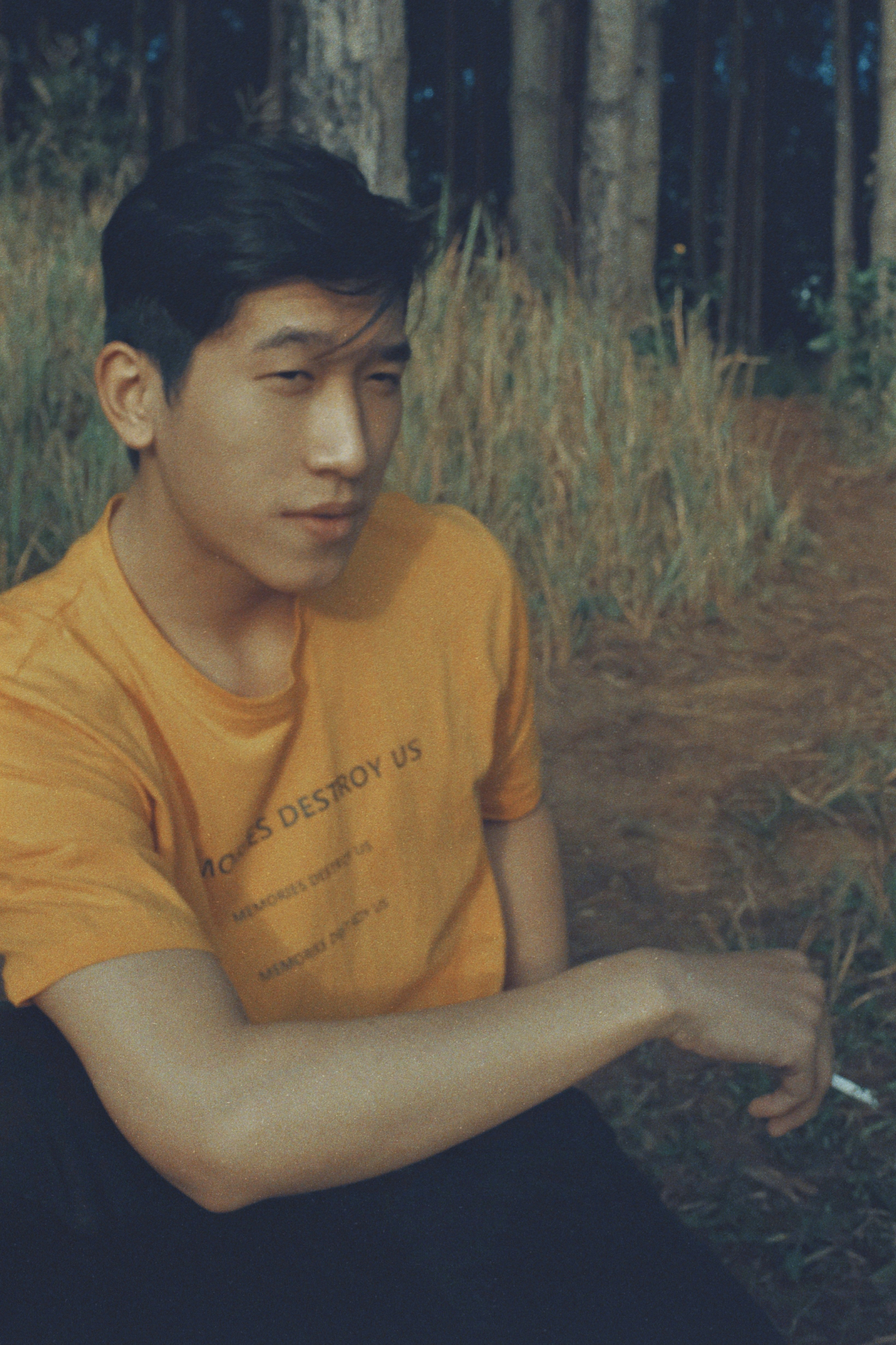 Young man in an orange shirt sitting amidst tall grass and trees, exuding a relaxed vibe.