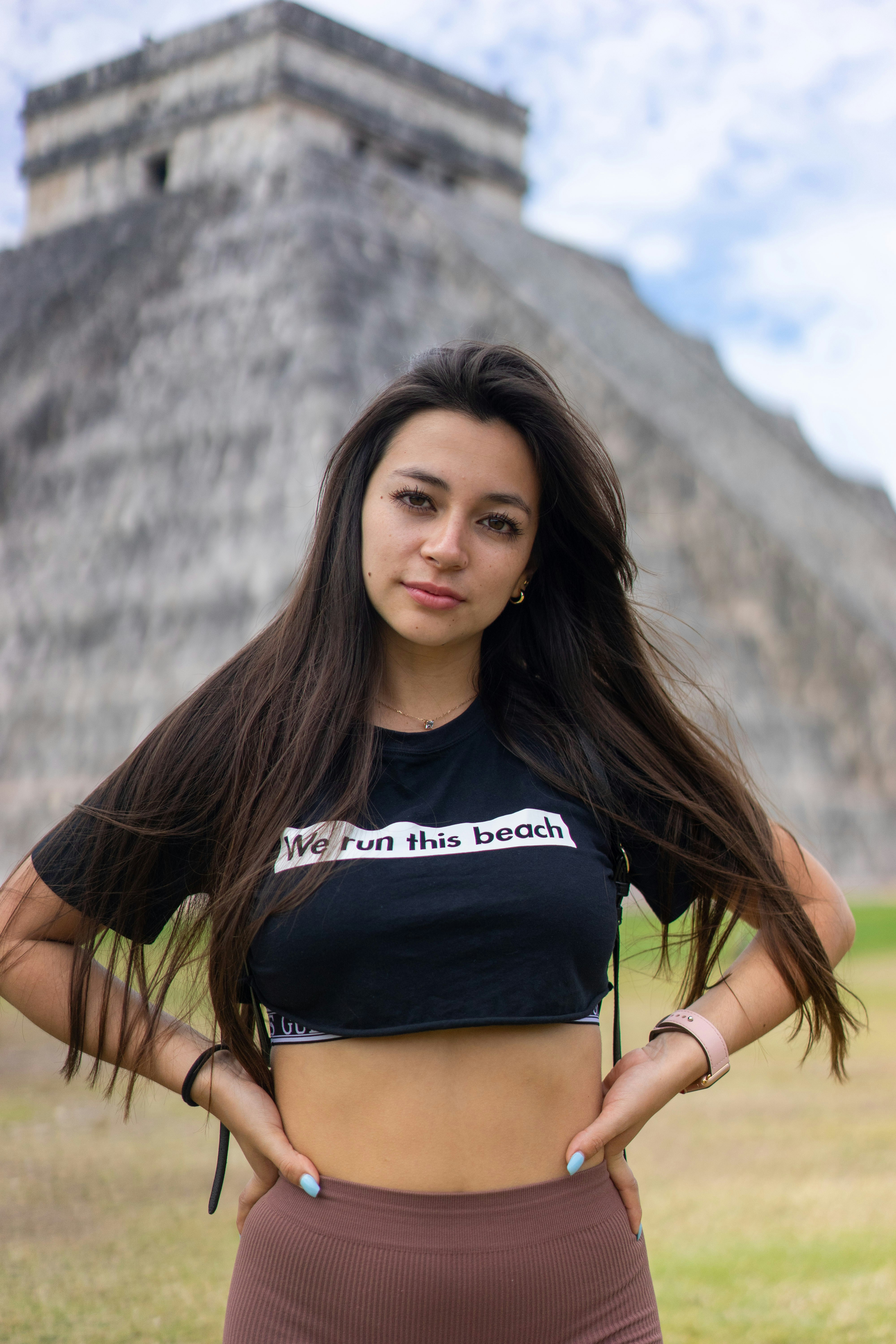 Young woman posing confidently in front of an ancient pyramid, showcasing a blend of contemporary fashion and historical backdrop.
