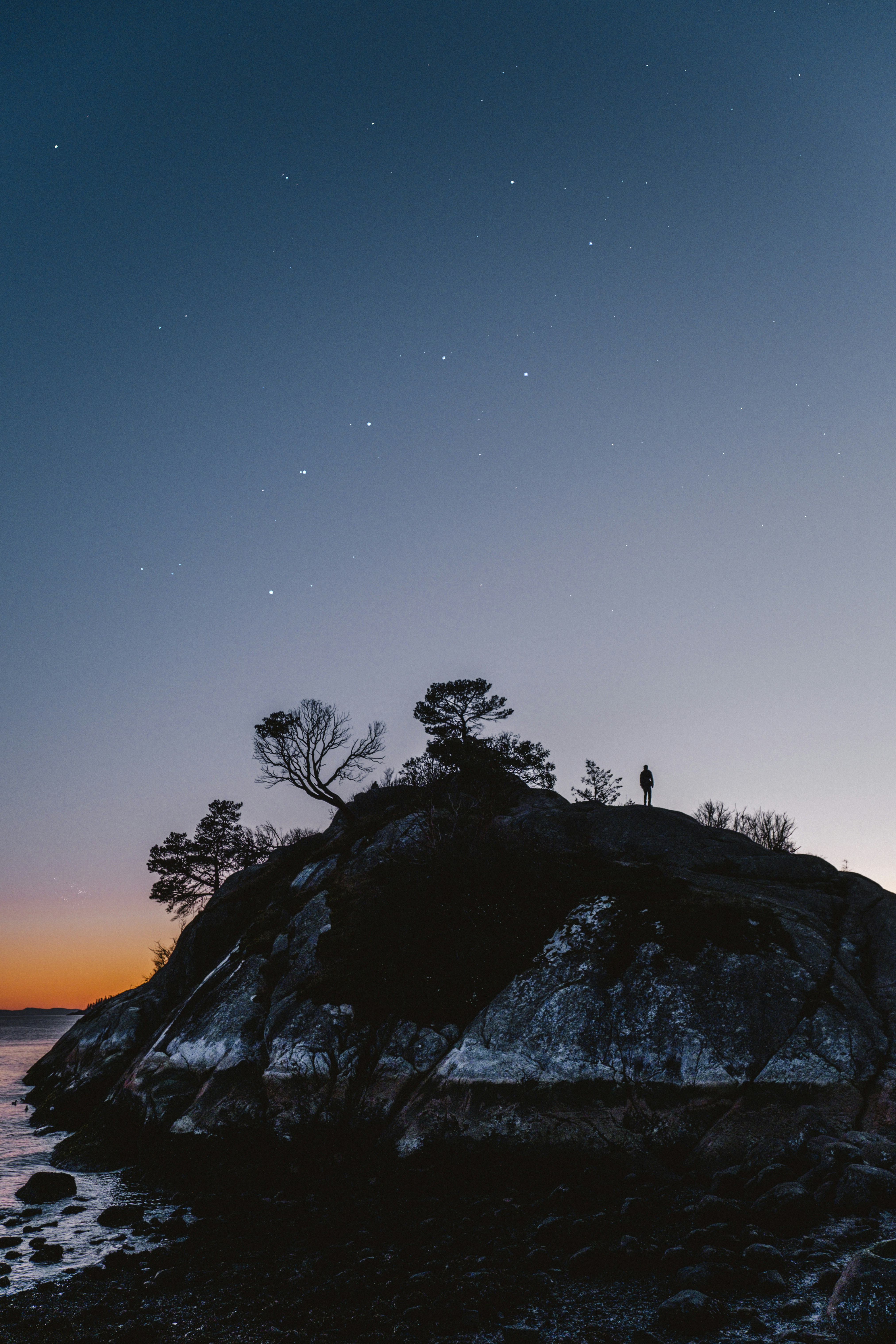 Photograph of a lone figure on a wind-sculpted rocky island beneath a gradient night sky dotted with stars.