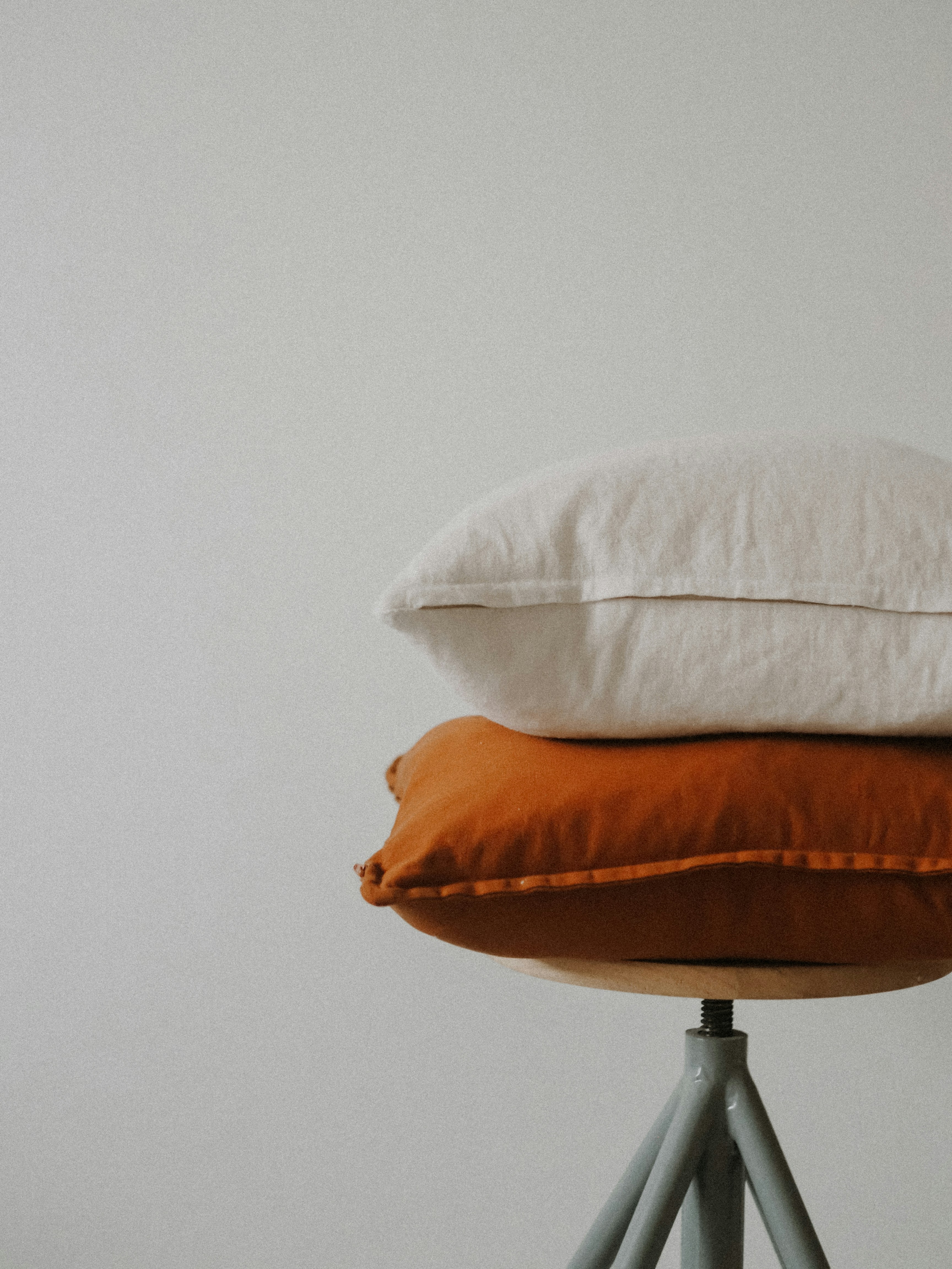 Two stacked pillows, one white and one terracotta, resting on a minimalist stool against a neutral wall.