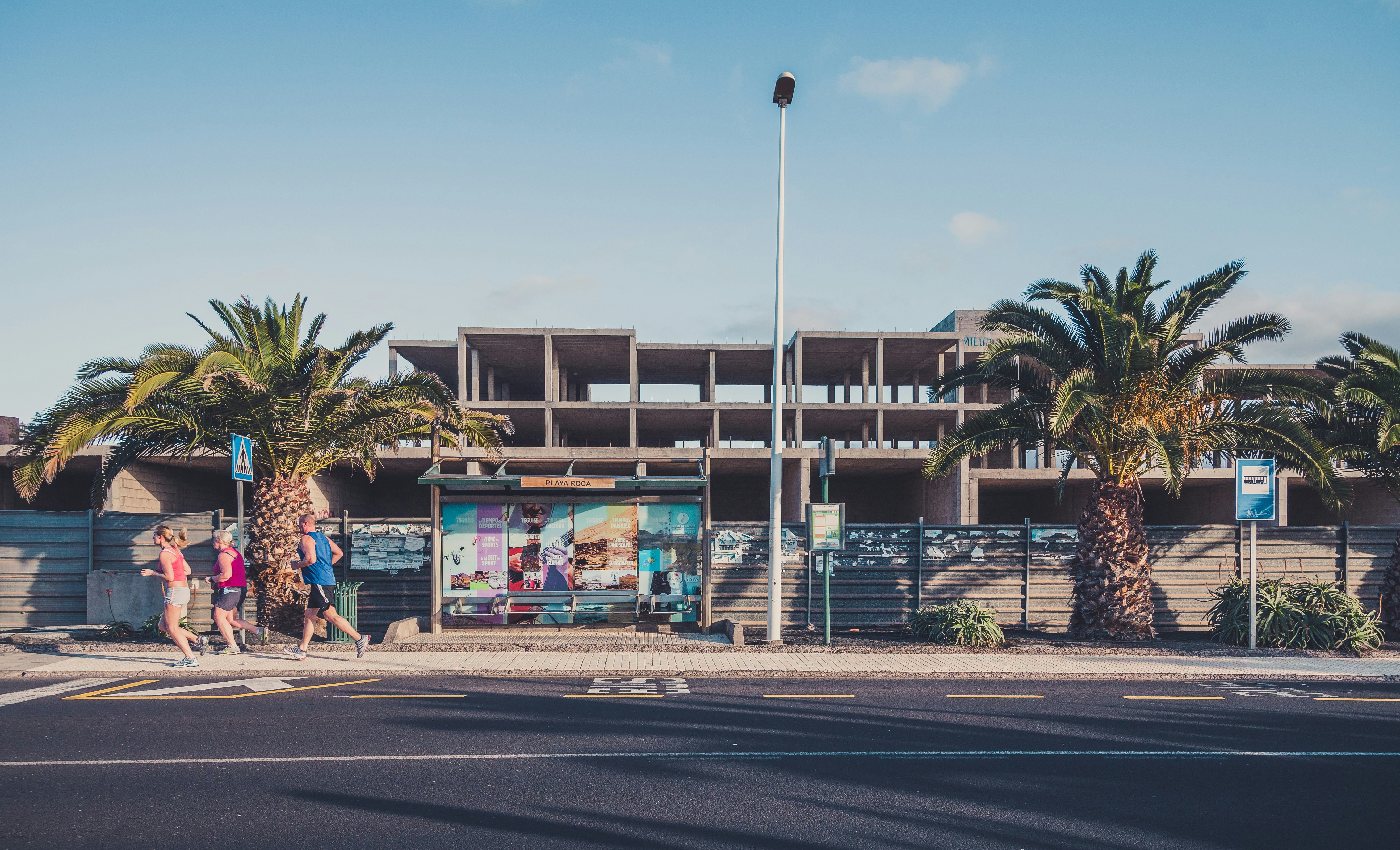 A vibrant scene featuring pedestrians walking past a bus stop framed by palm trees, with a partially constructed building in the background. The image captures the essence of urban life interwoven with natural elements.