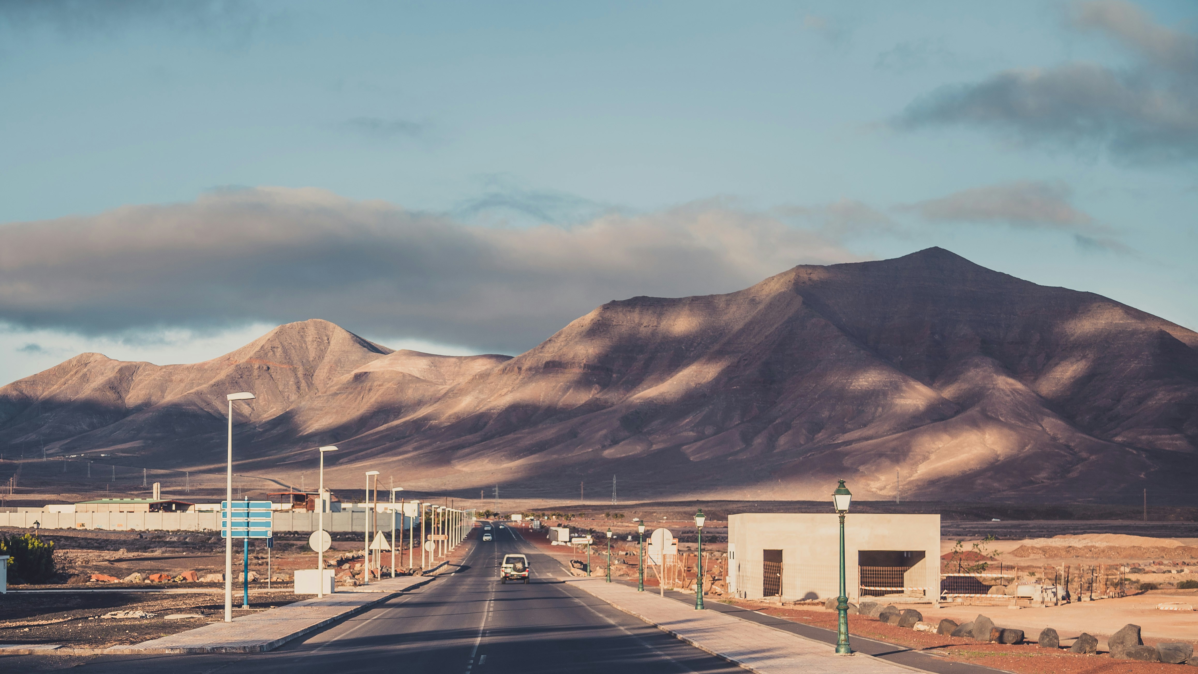 A winding road leads through a vast desert landscape, flanked by towering mountains casting long shadows under a clear sky.