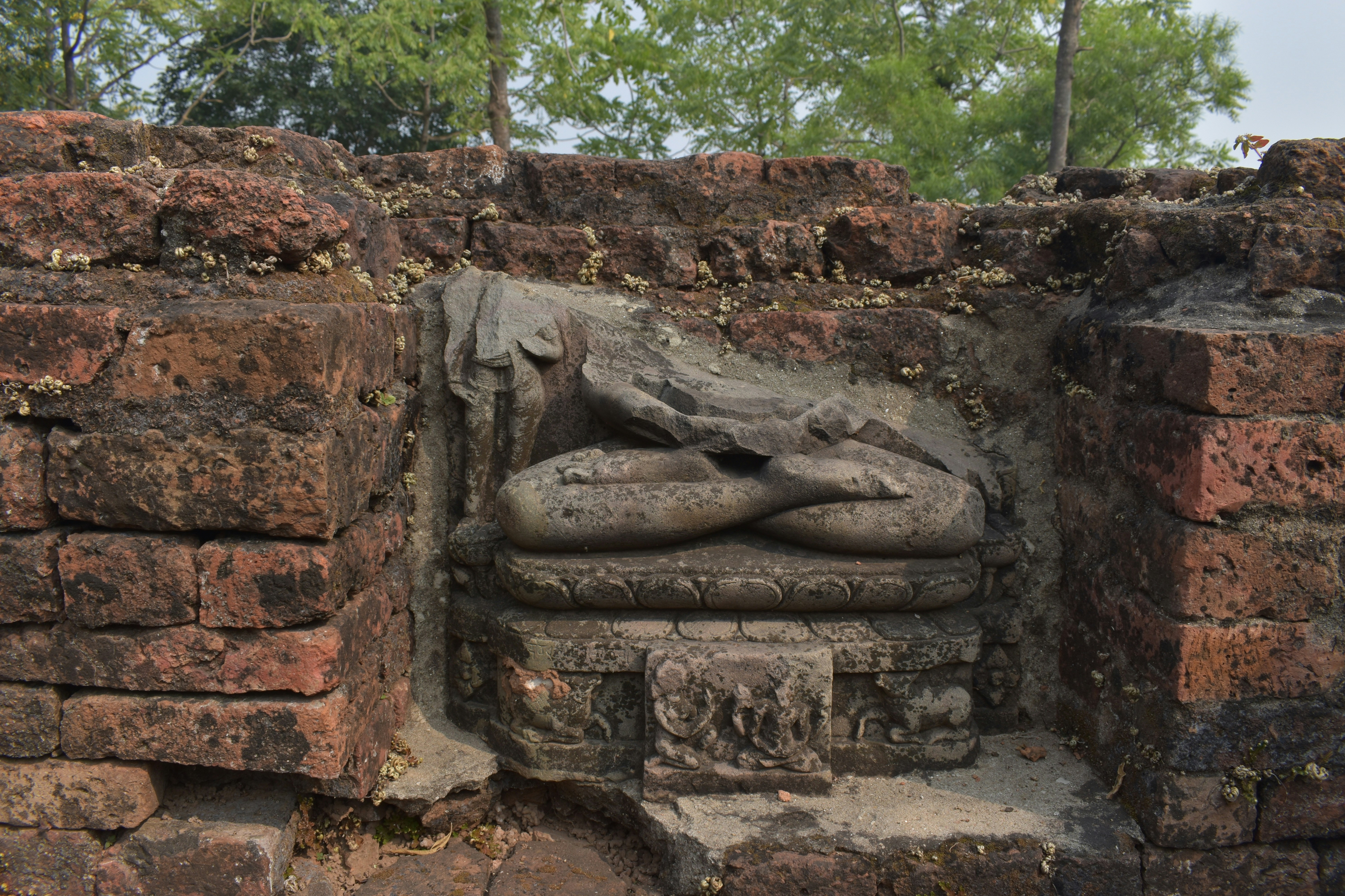Weathered stone statue embedded in a brick wall, framed by lush green trees.