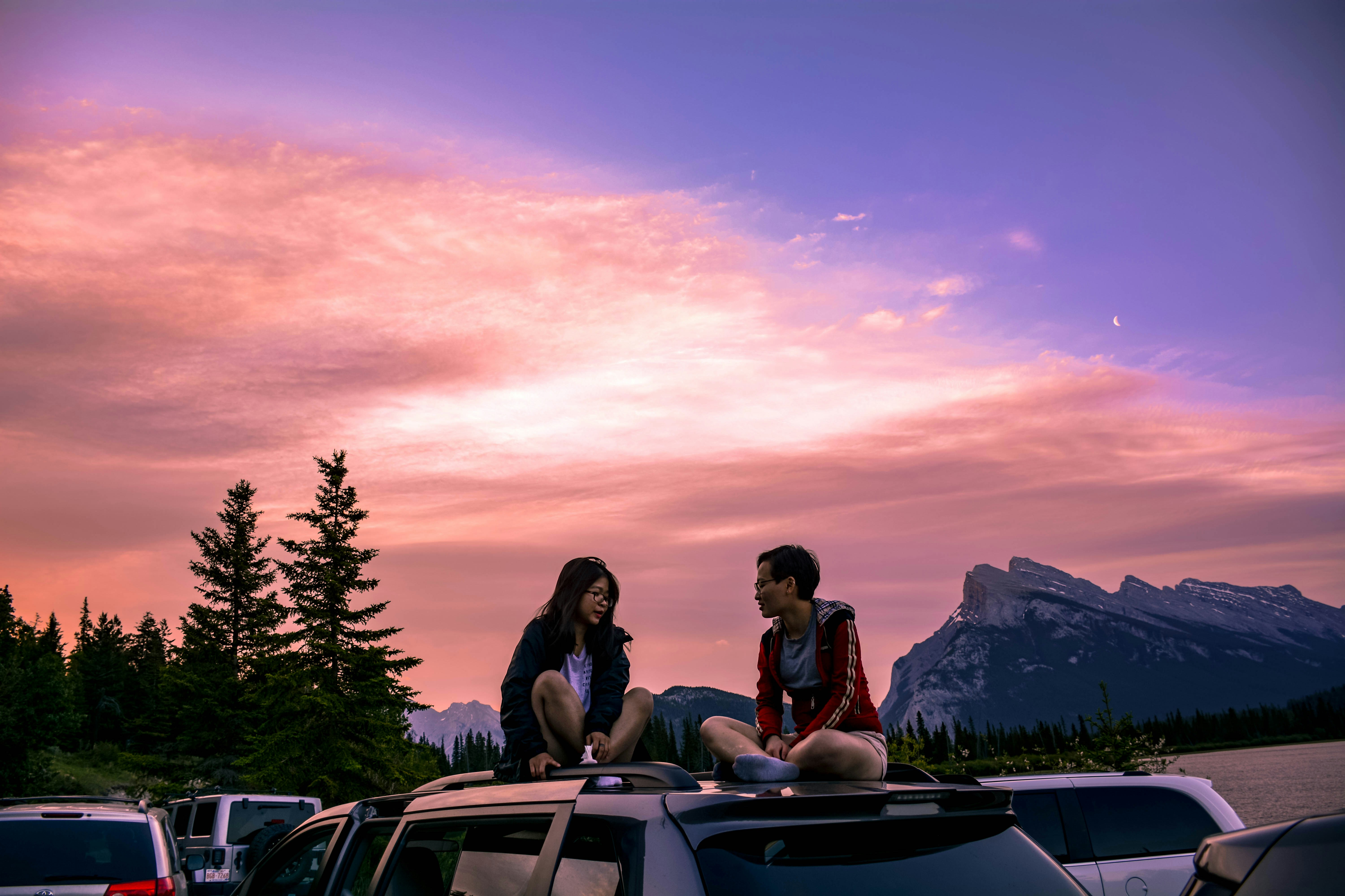 Two people sitting on a car roof under a vibrant sunrise sky with mountains in the background.