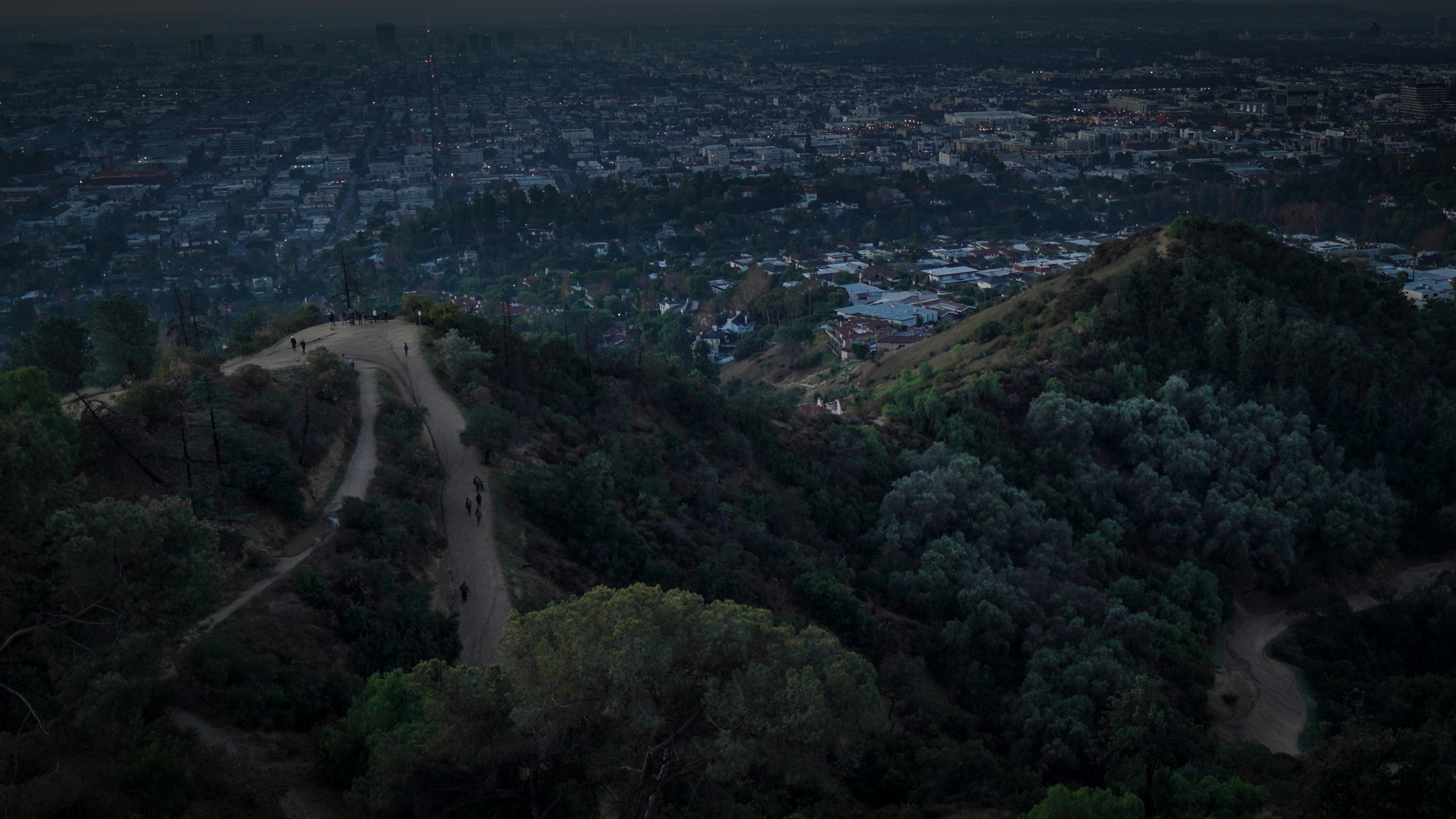 A winding path through a hilly landscape, overlooking a sprawling city at twilight. The scene captures the transition from nature to urban life.