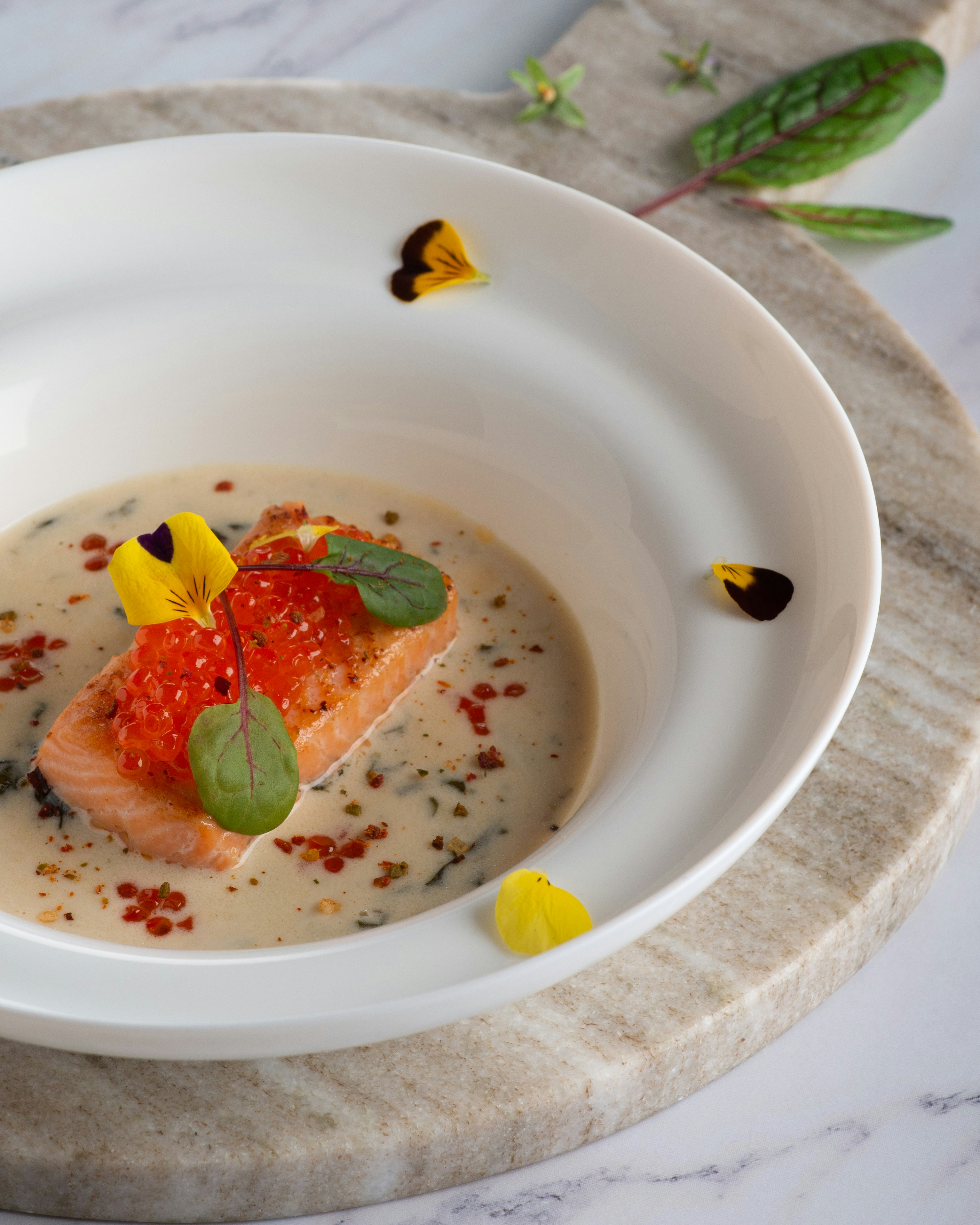 A beautifully plated salmon fillet garnished with vibrant red caviar and delicate edible flowers, resting in a creamy sauce. The dish is presented on a marble surface, enhancing its gourmet appeal.