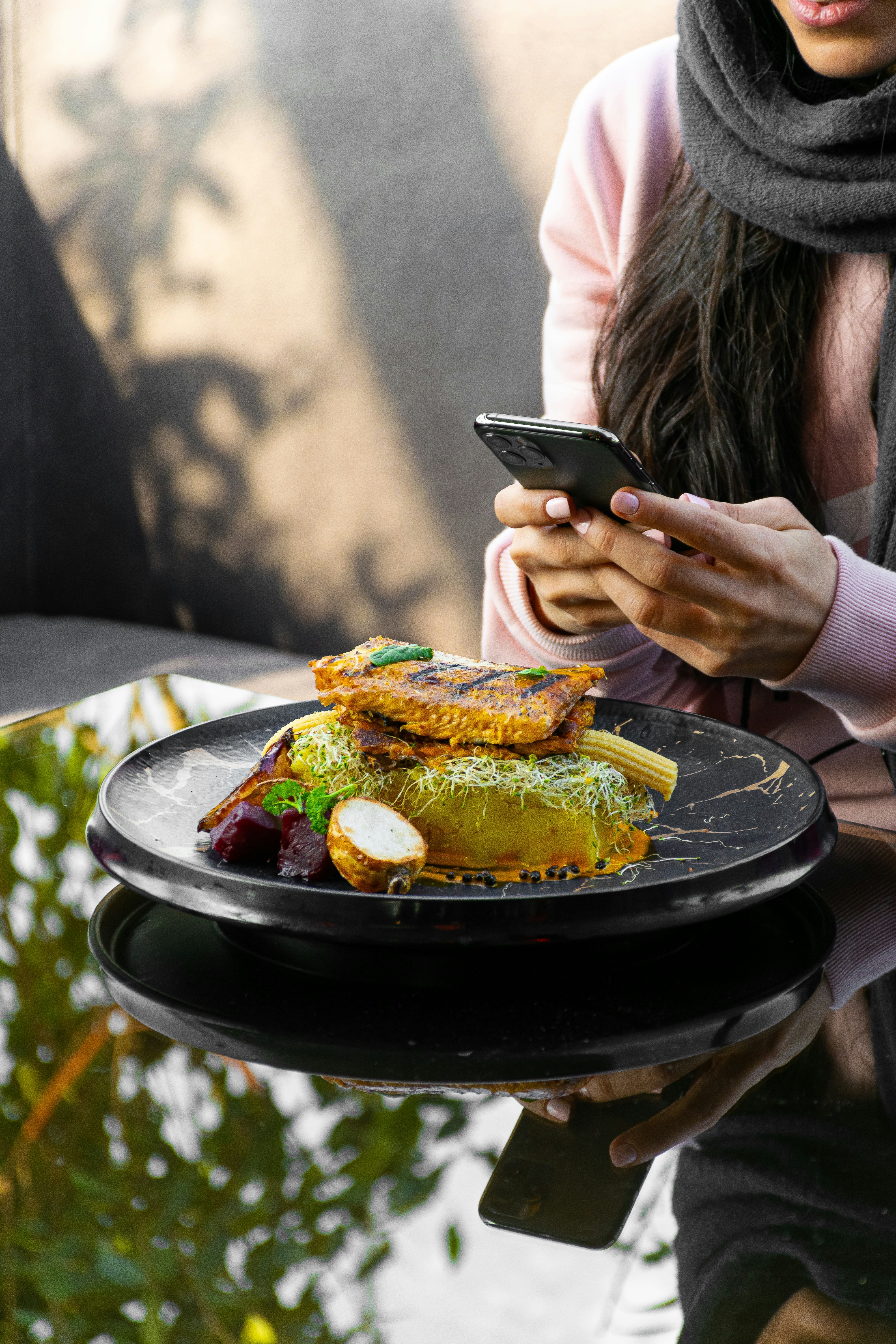 Gourmet dish featuring grilled protein, vibrant vegetables, and artistic garnishes, served on a sleek black plate. A diner captures the moment with a smartphone.