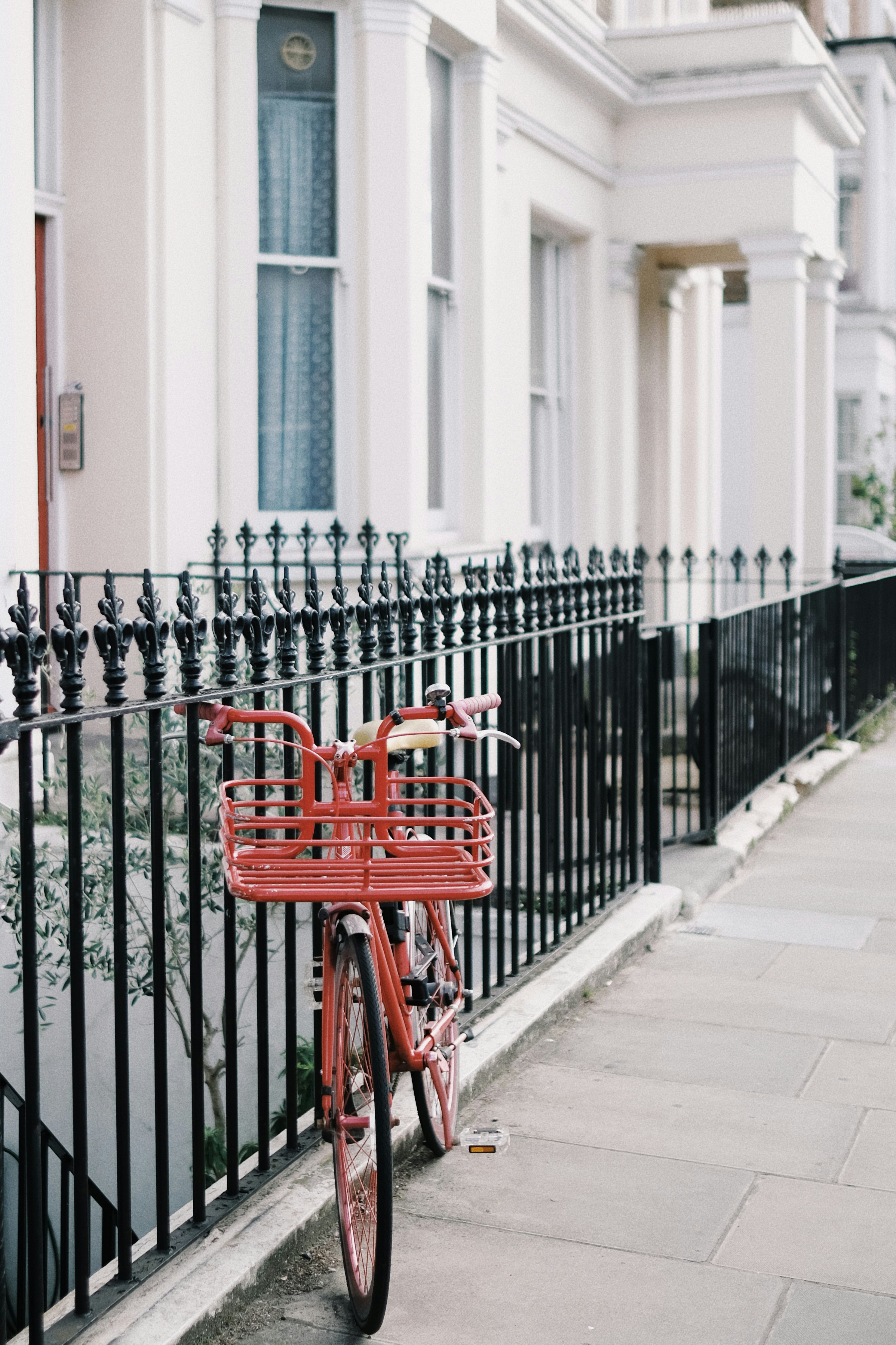 A vibrant red bicycle rests against a black wrought iron fence in a quaint urban setting. The scene captures a blend of architectural elegance and everyday life.