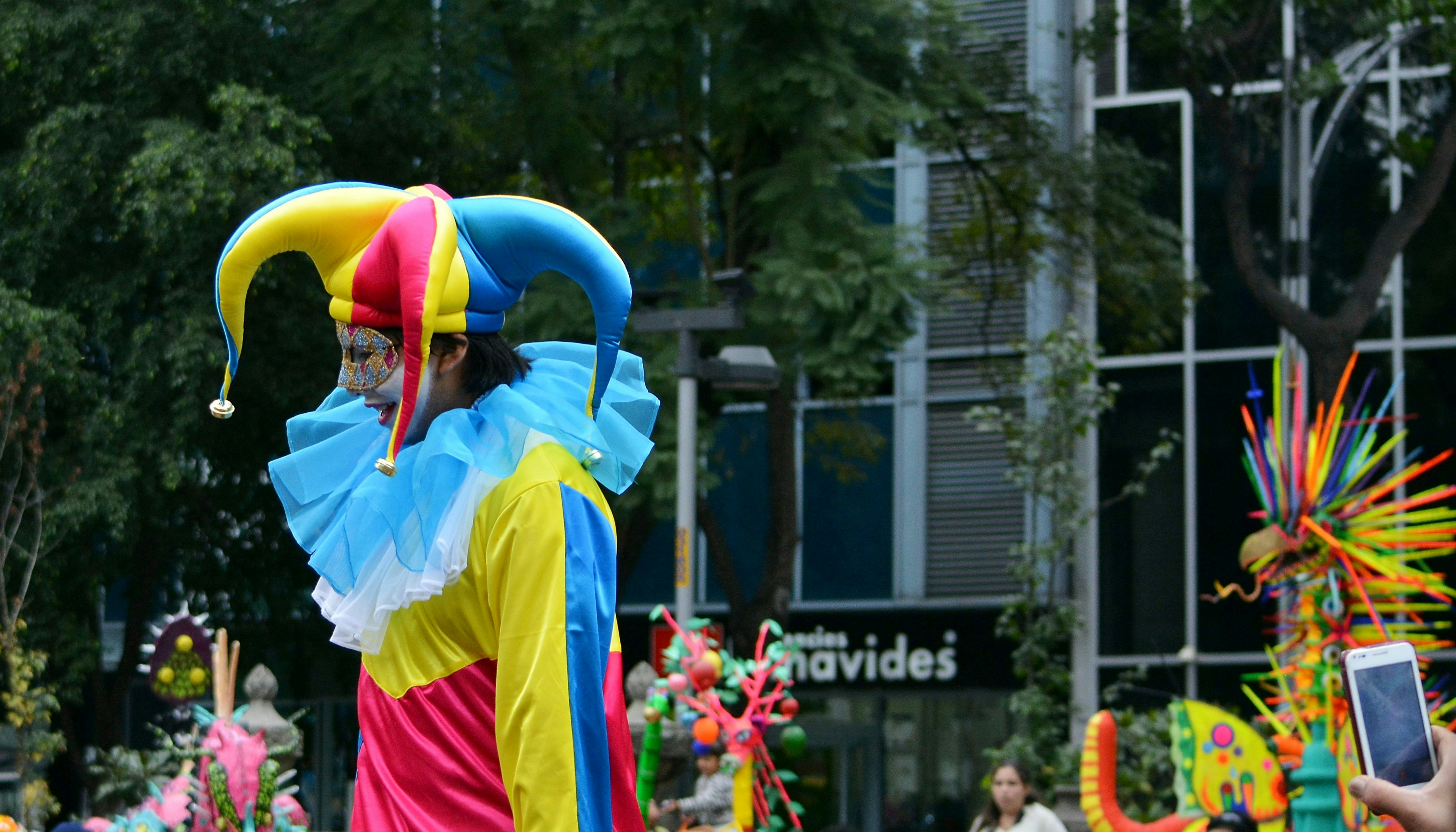Colorful street parade photograph featuring a jester in blue, yellow, and pink with a voluminous collar, with vivid parade floats in the background.