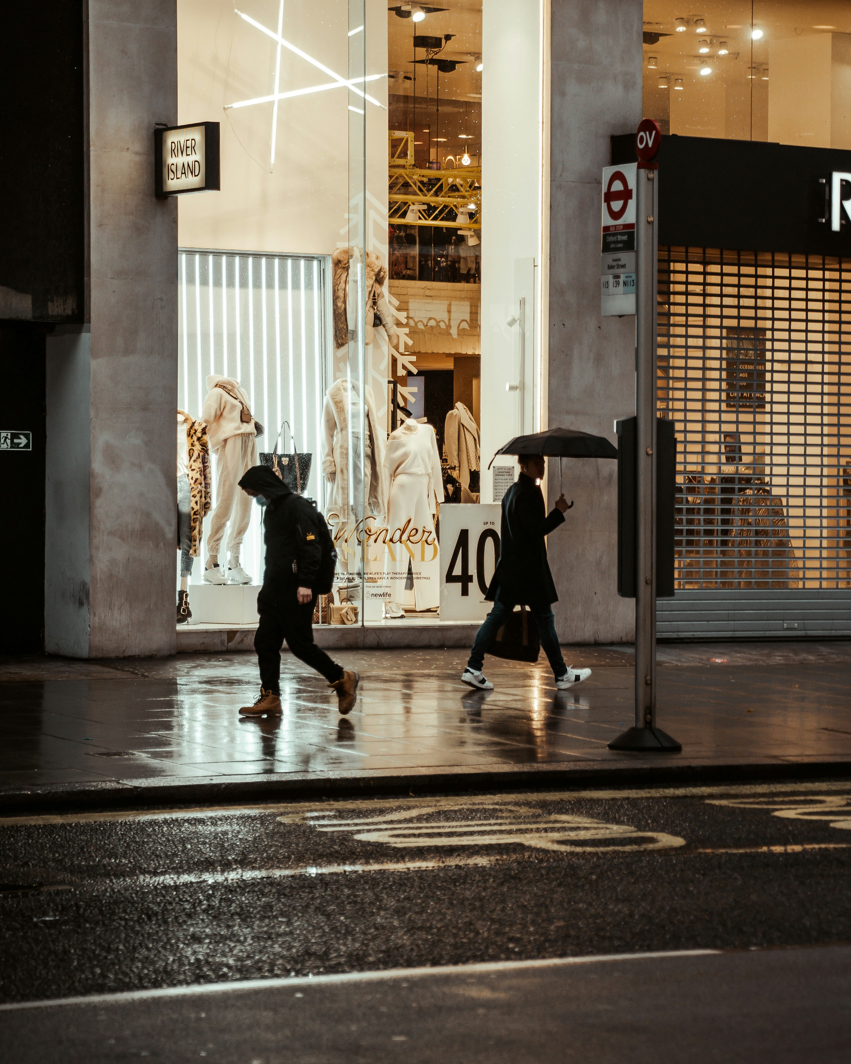 Two pedestrians navigate a wet street under umbrellas, passing by a fashion store's illuminated window display. The scene captures the essence of urban life on a rainy evening.