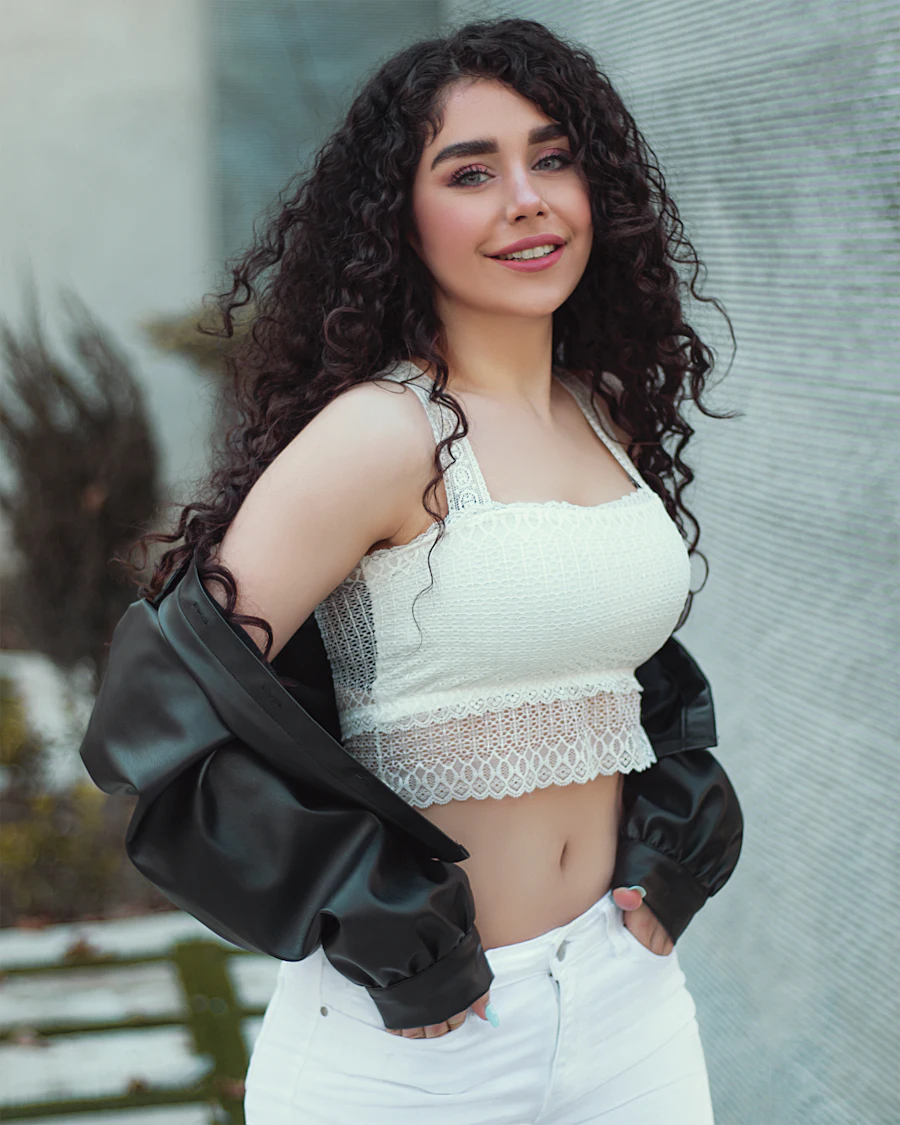 Smiling woman with voluminous curly hair posing in a white lace crop top and black jacket, standing against a textured outdoor wall. Soft background blur keeps attention on her face.