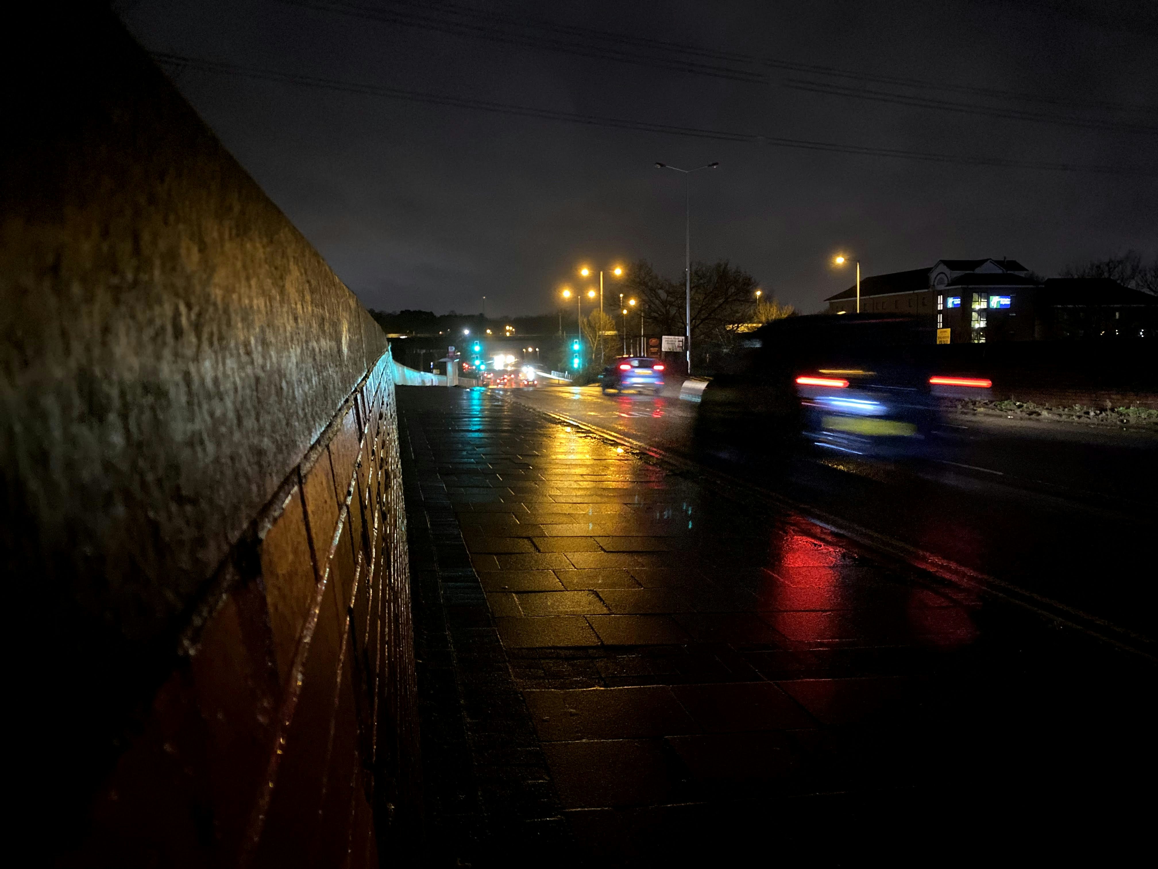 Street scene with blurred car lights reflecting on wet pavement under a dark sky.