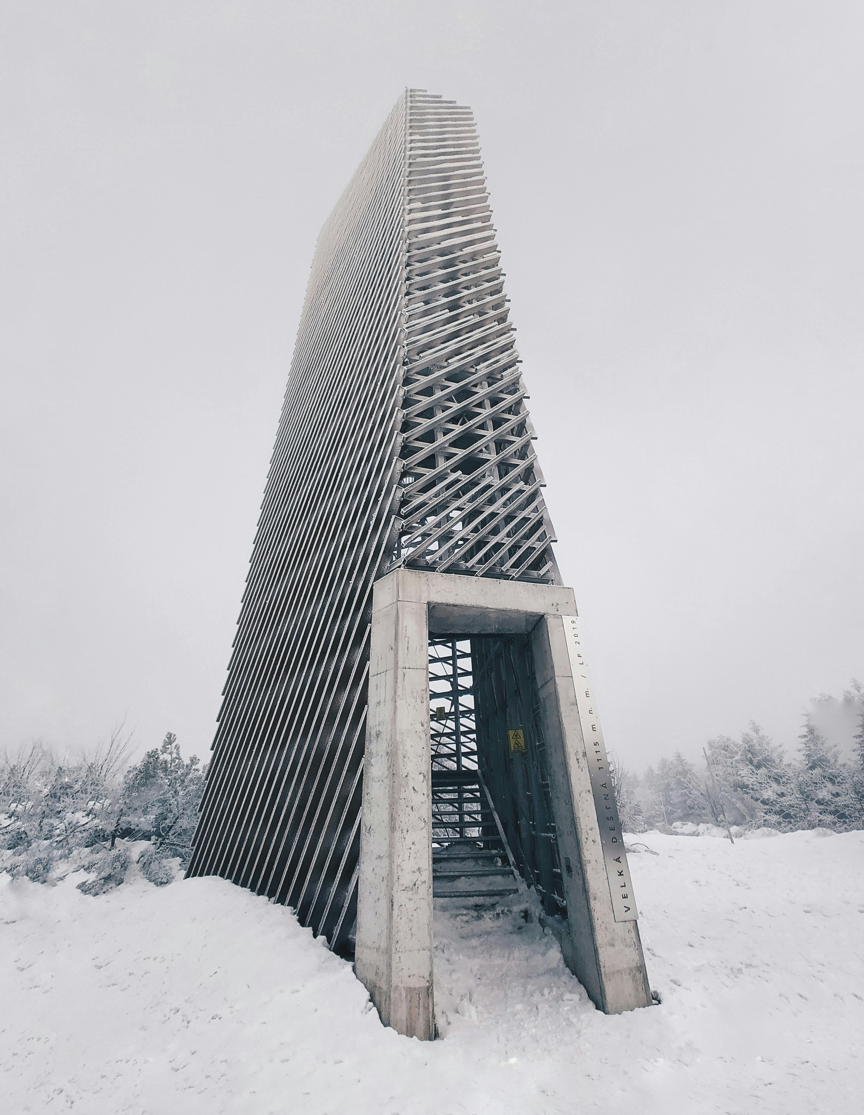 Tall, modern wooden tower rising from a snowy landscape under a grey sky. The architectural design contrasts with the surrounding winter scenery.