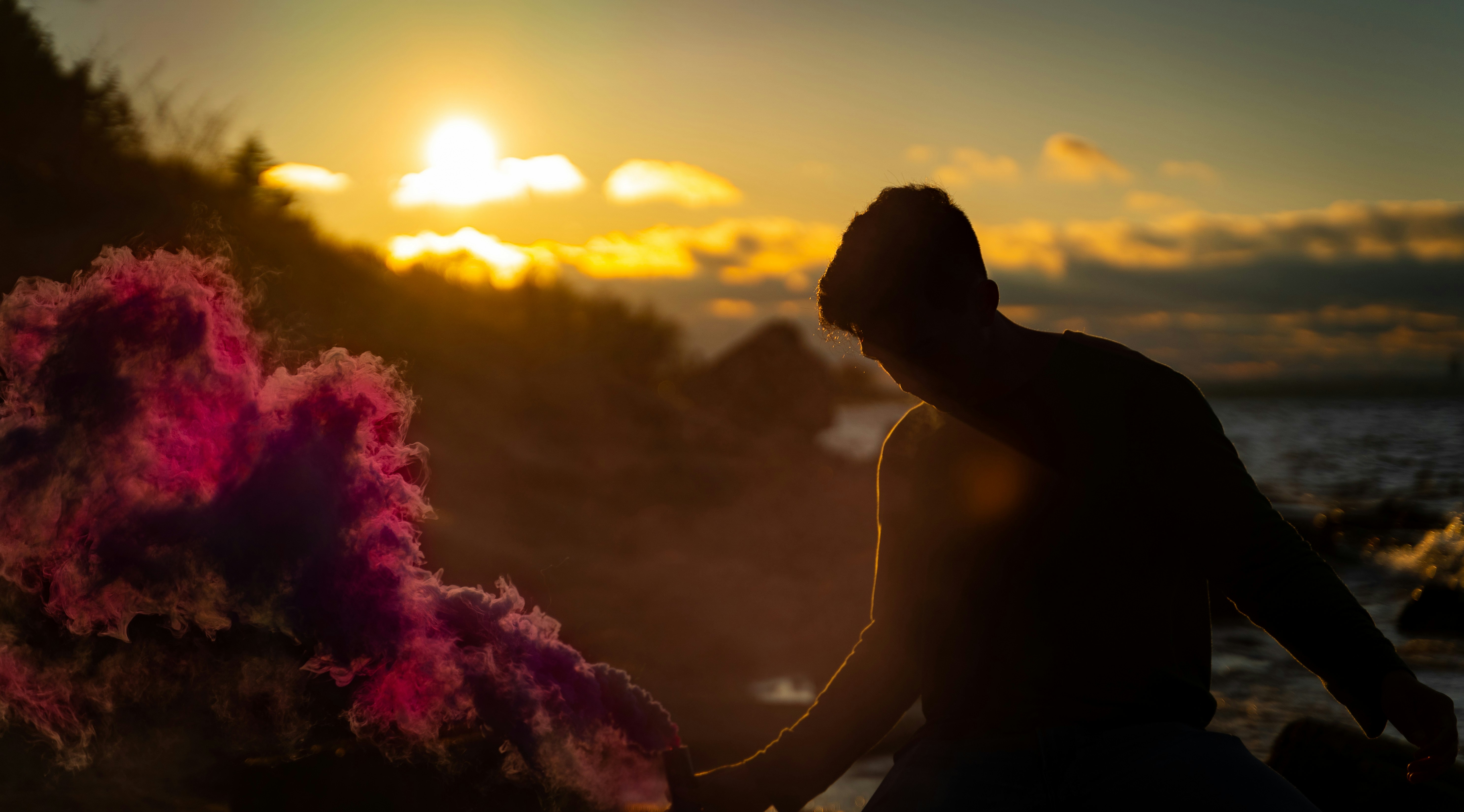 Silhouette of a person with vibrant purple smoke against a sunset sky by the shoreline.