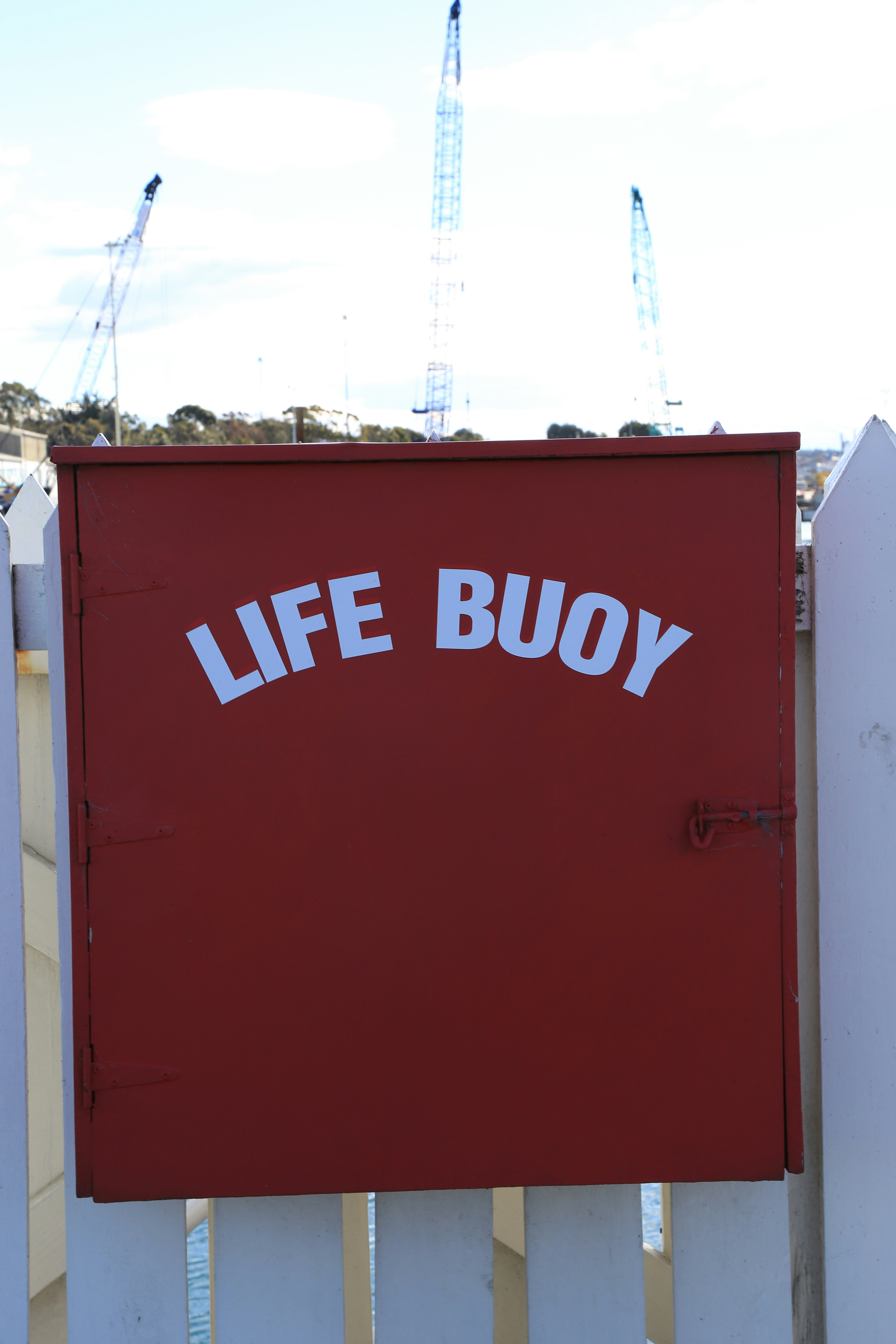 Bright red life buoy storage box against a backdrop of cranes and water. The box prominently displays 'LIFE BUOY' in white lettering.