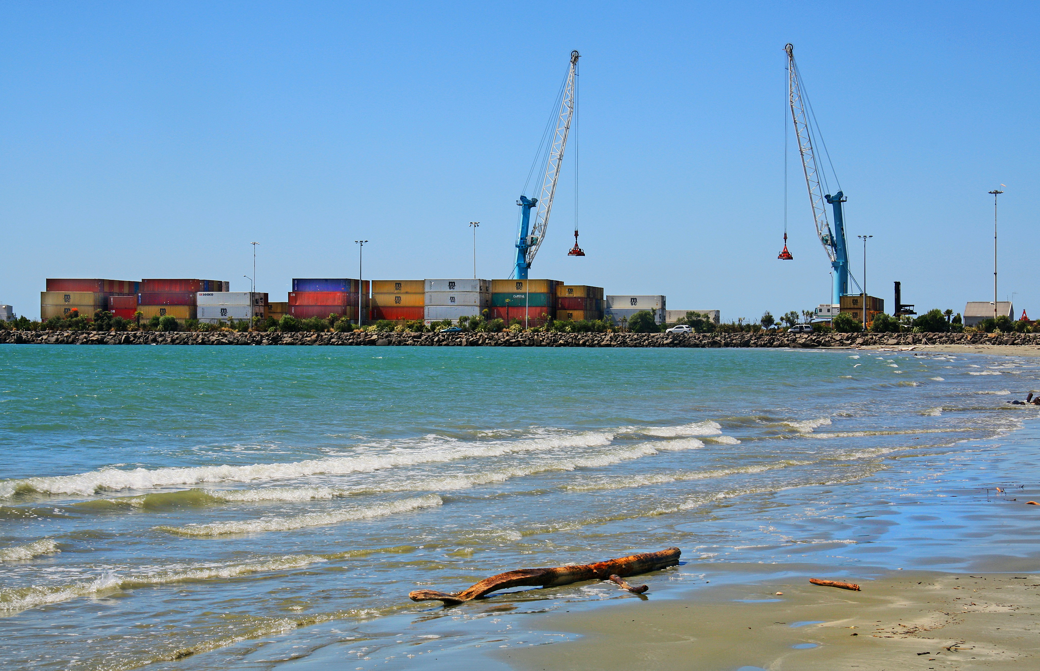Cranes work on stacked containers in the distance, In front is a rocky breakwater and a gently curving beach with white foam and driftwood logs.