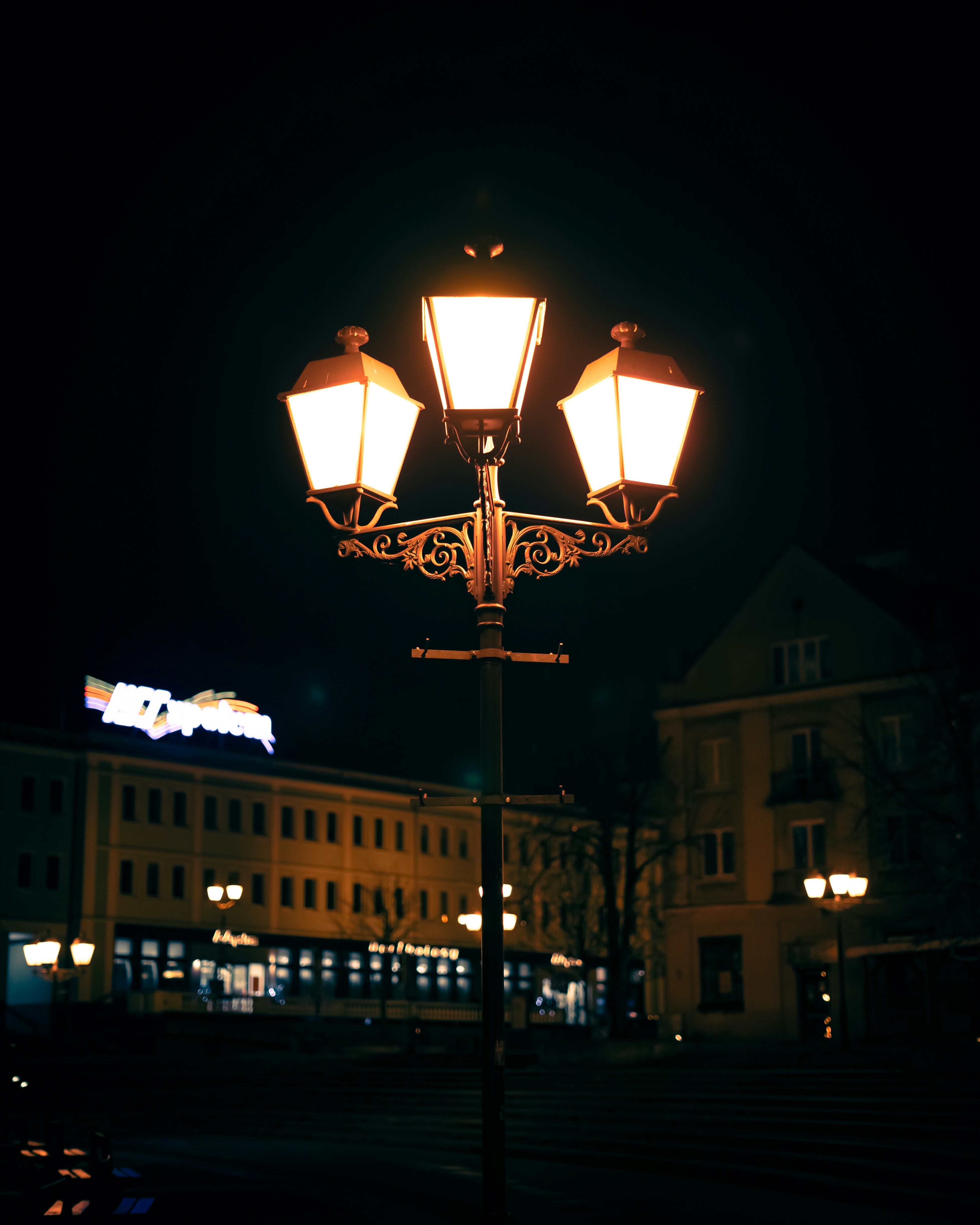 Vintage street lamp illuminating a nighttime square, surrounded by softly glowing lights from nearby buildings.