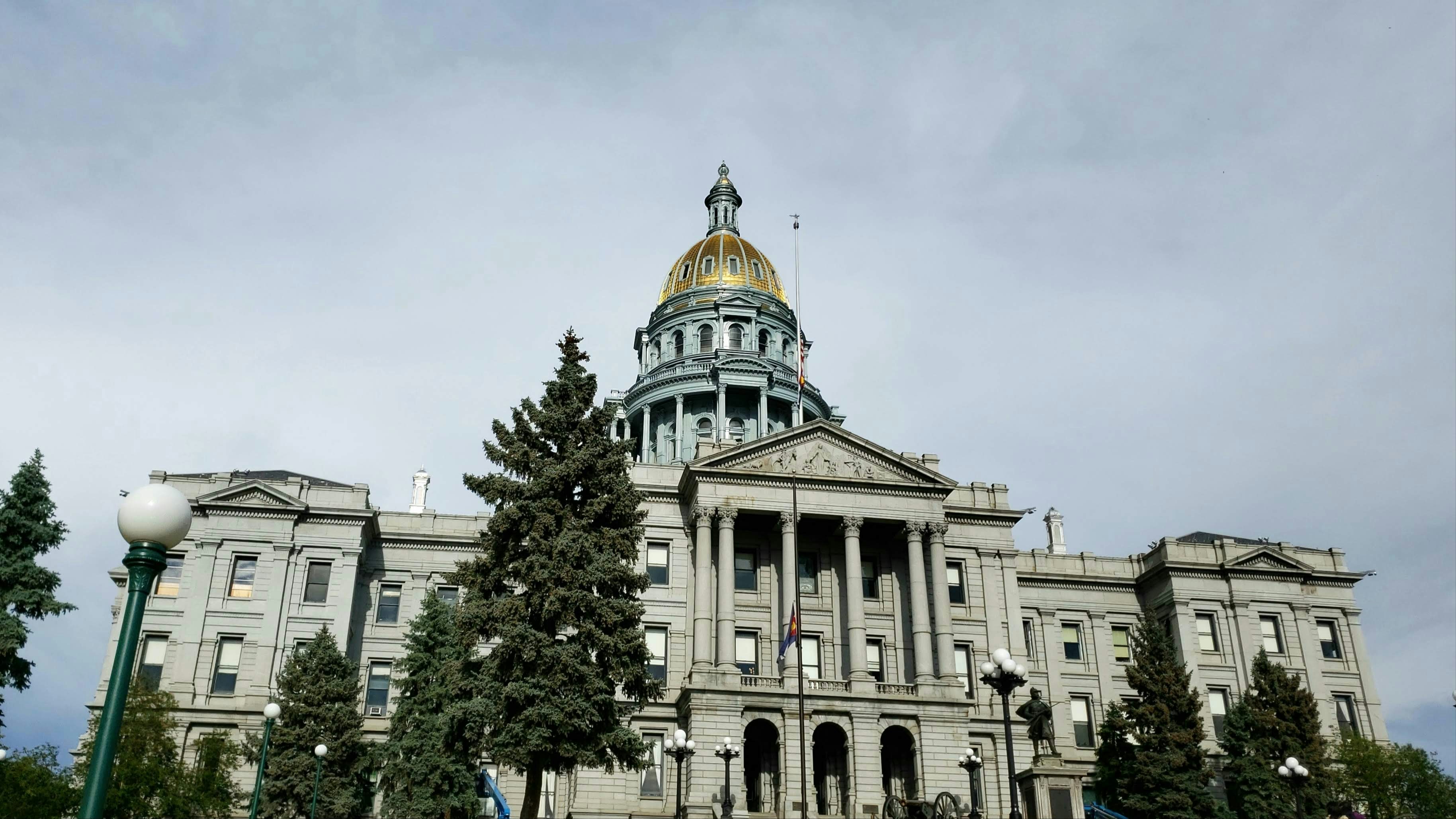 Historic capitol building with a golden dome surrounded by tall trees under a cloudy sky.