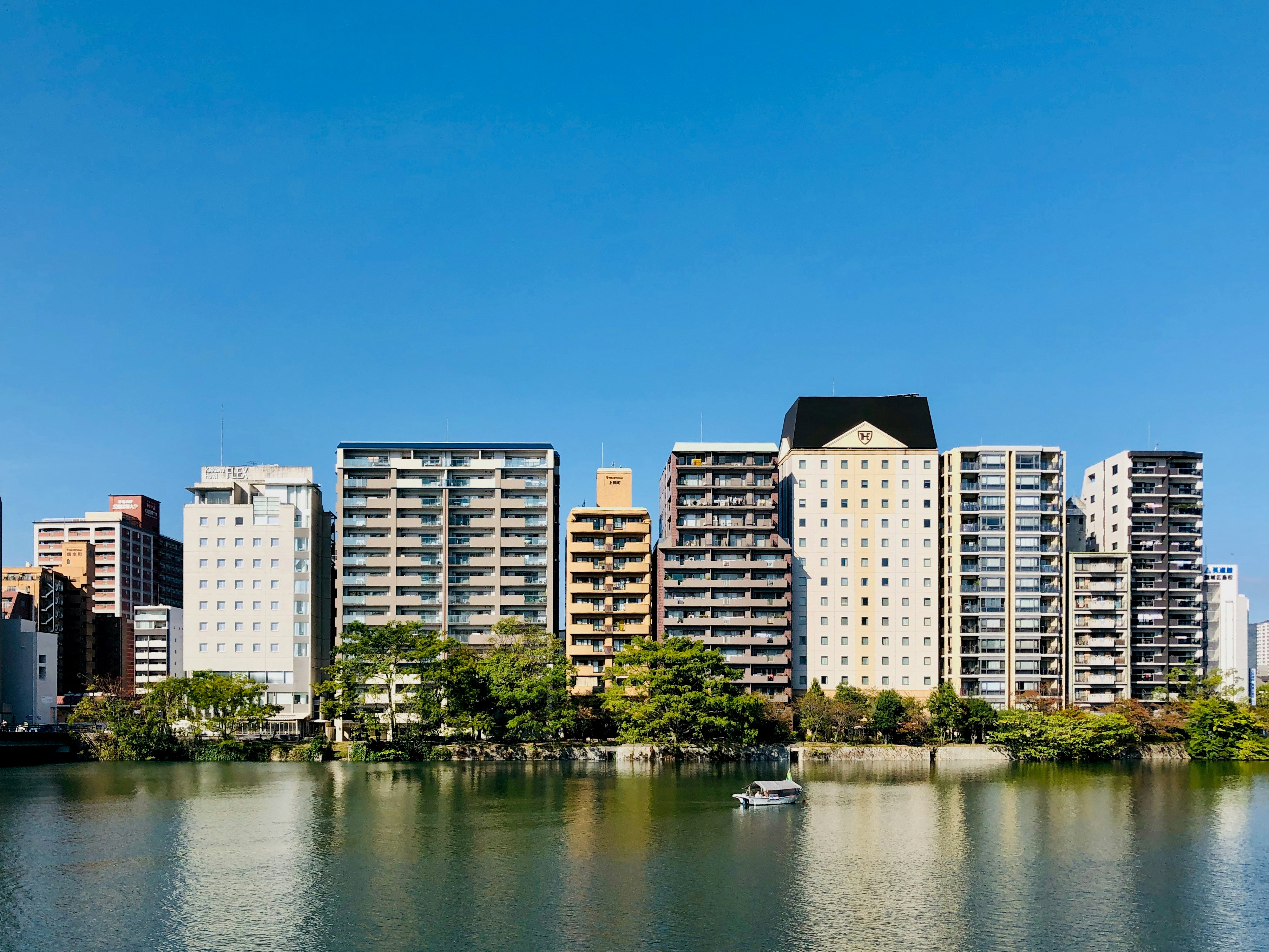 City skyline with diverse architecture mirrored in a calm body of water under a clear blue sky.