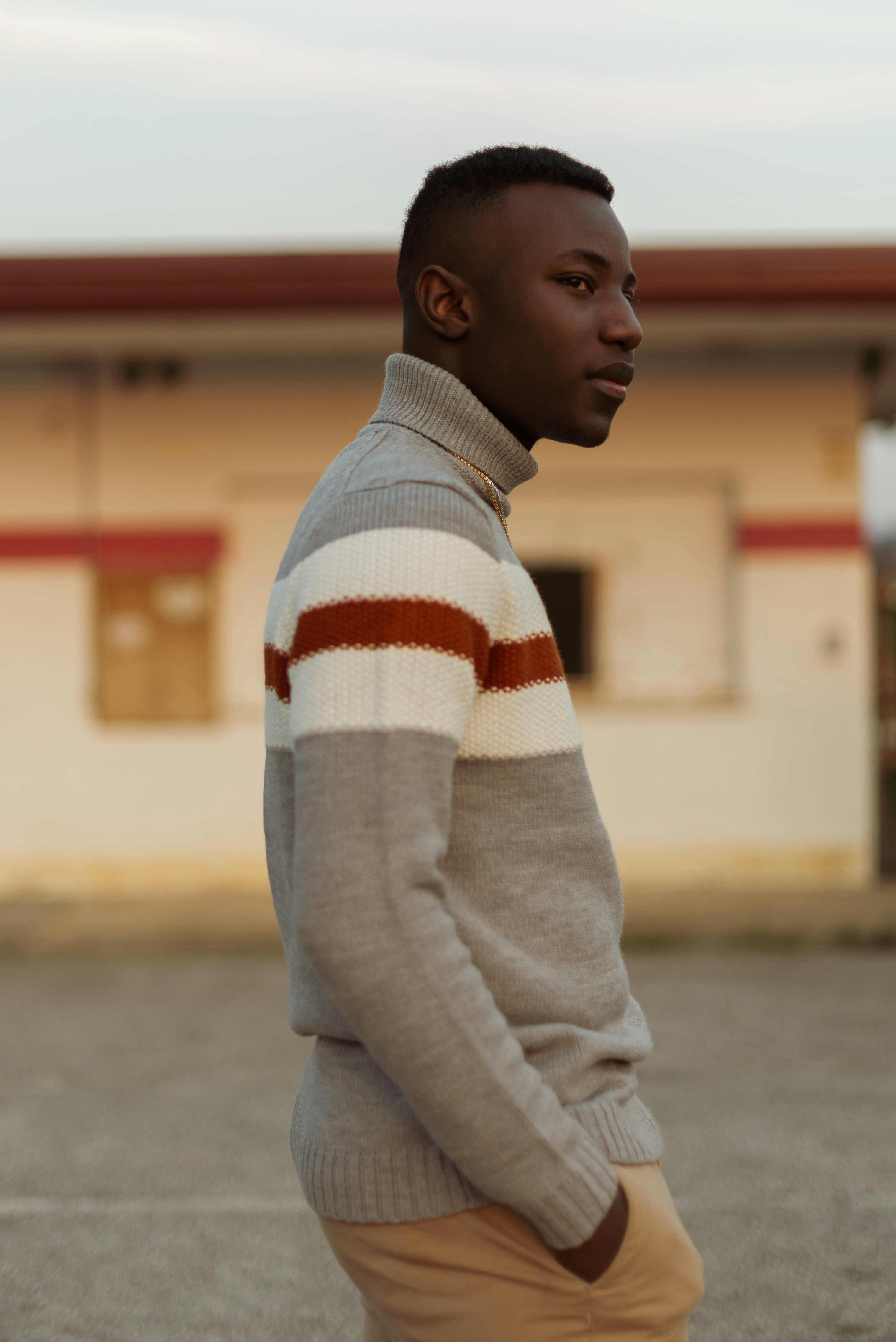 Young man in a stylish sweater gazing thoughtfully, with an urban backdrop softly blurred. His profile captures a moment of reflection.