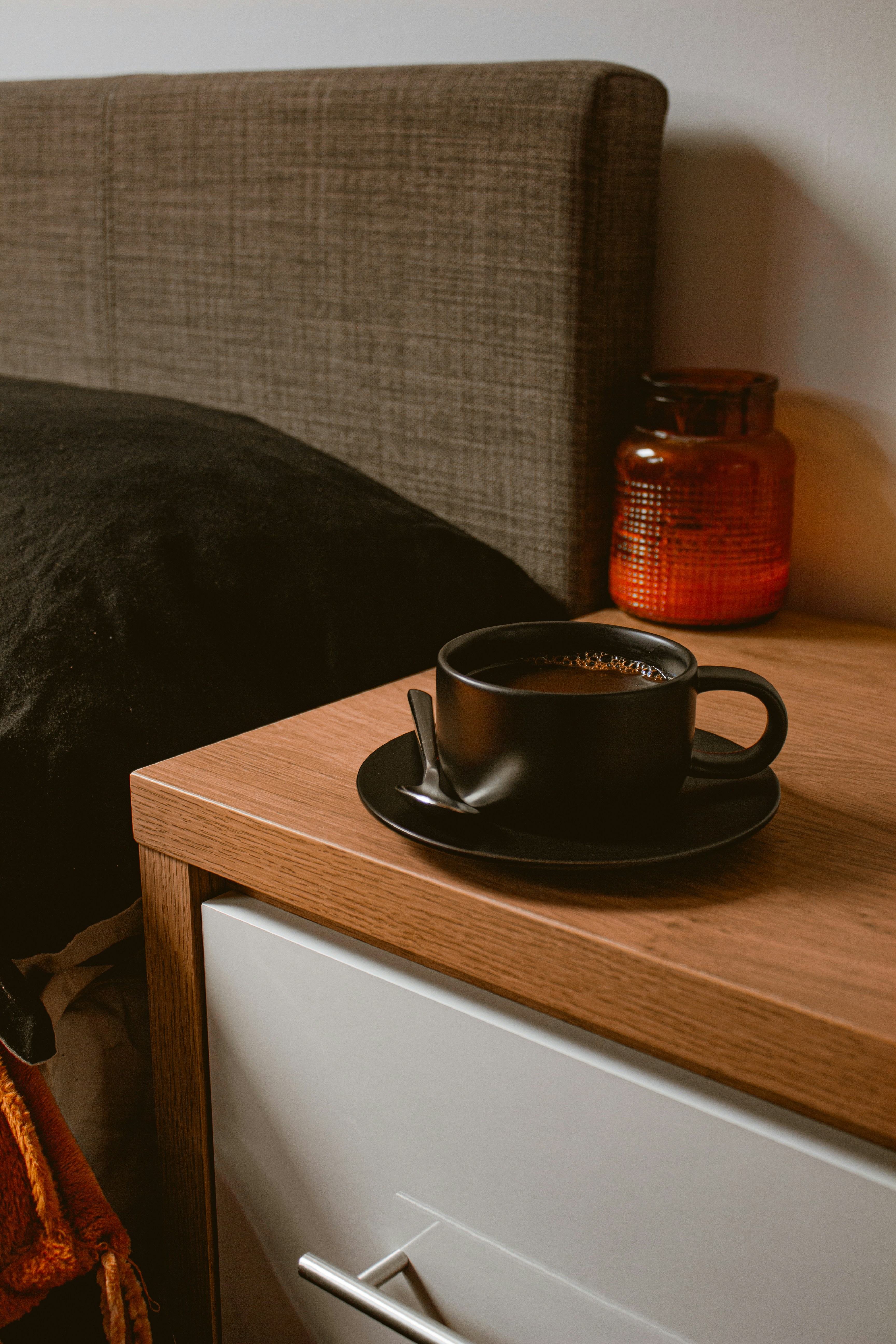 A black coffee cup rests on a wooden nightstand beside a cozy bed, complemented by a warm orange throw and a glowing glass jar.