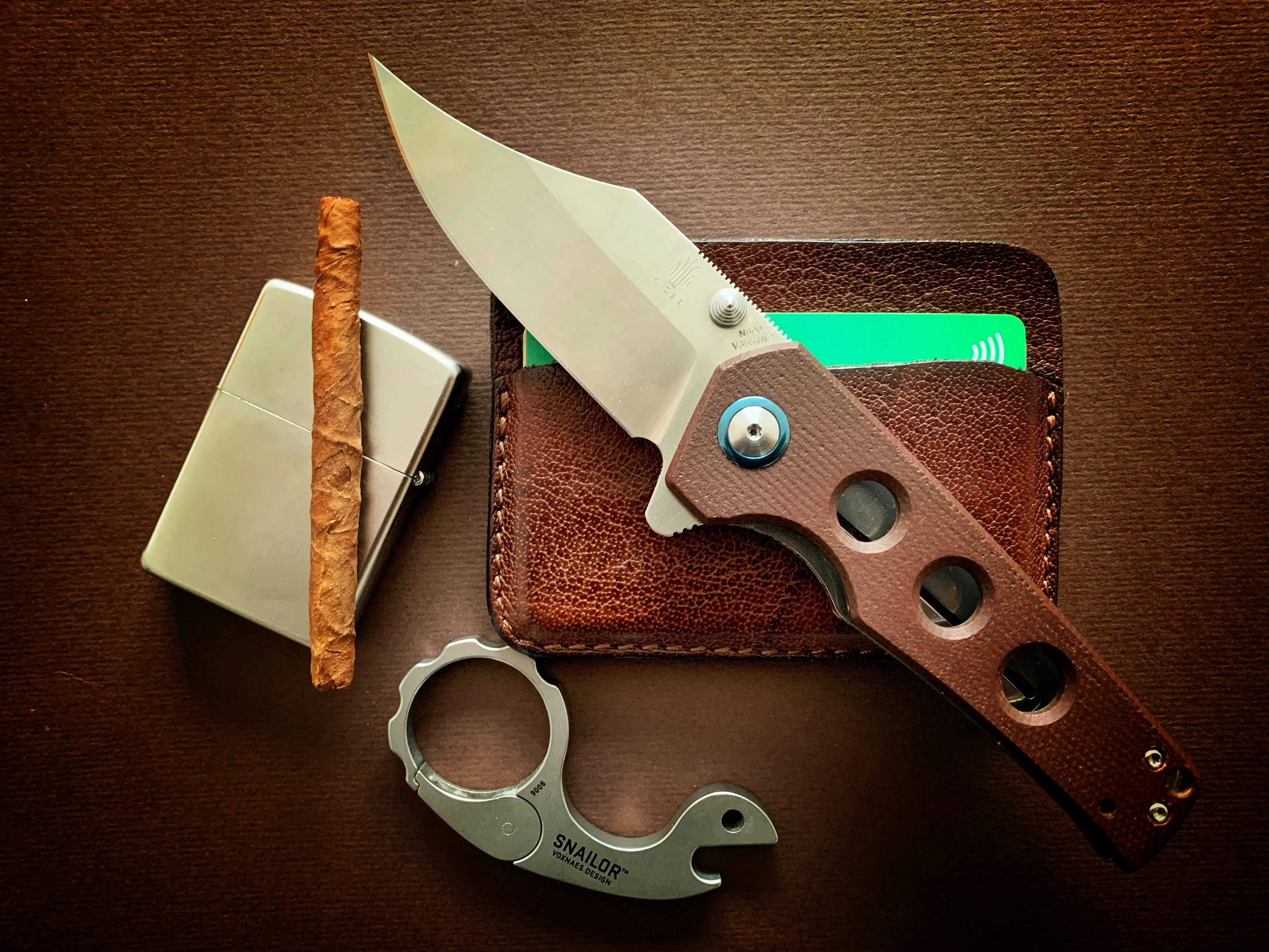 A sleek folding knife, a cigar, a lighter, and a multi-tool rest on a leather wallet, showcasing essential gear for outdoor enthusiasts.