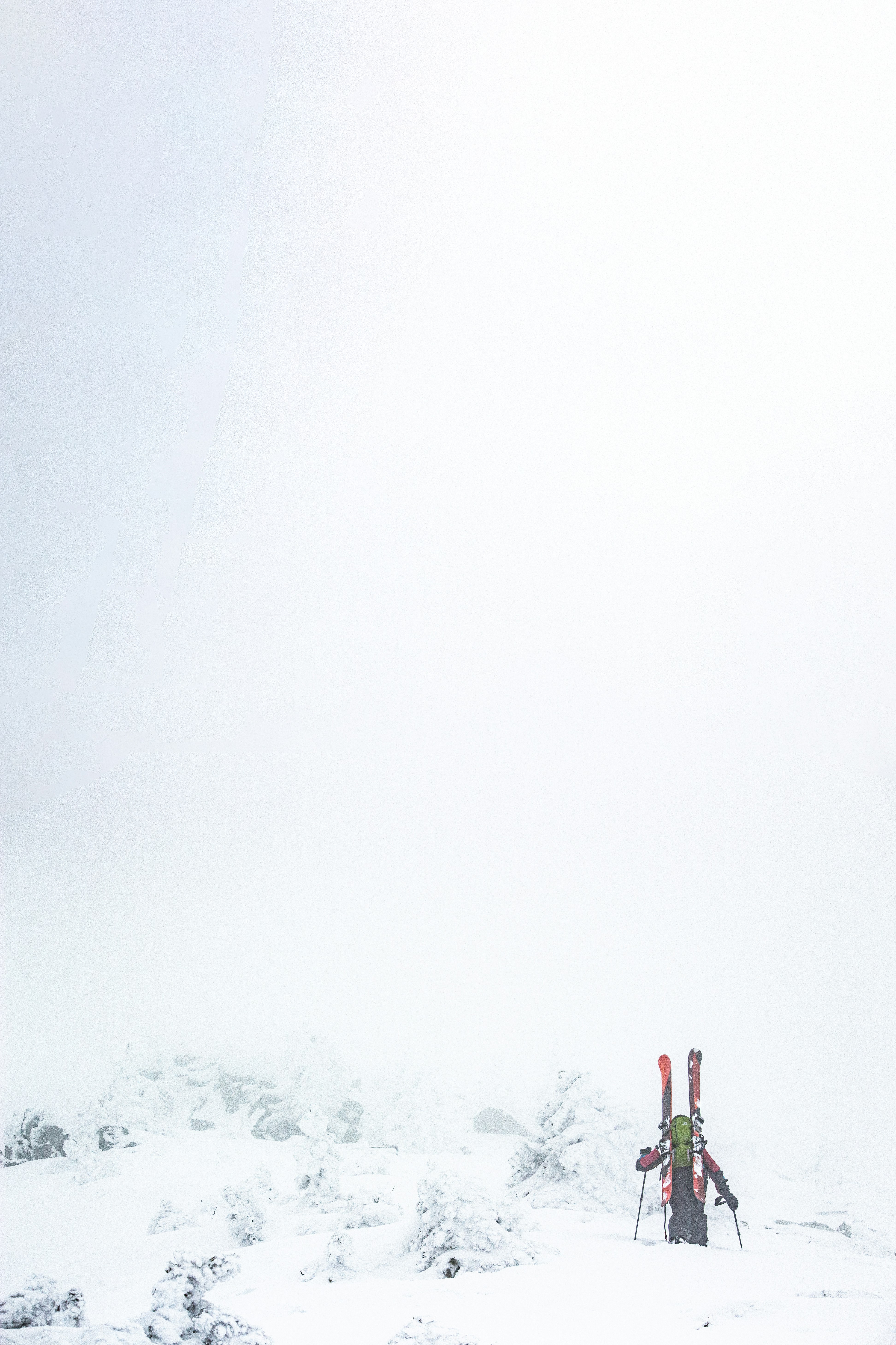 Skier standing in a snowy landscape shrouded in fog, holding ski poles with bright red skis resting on their shoulders.