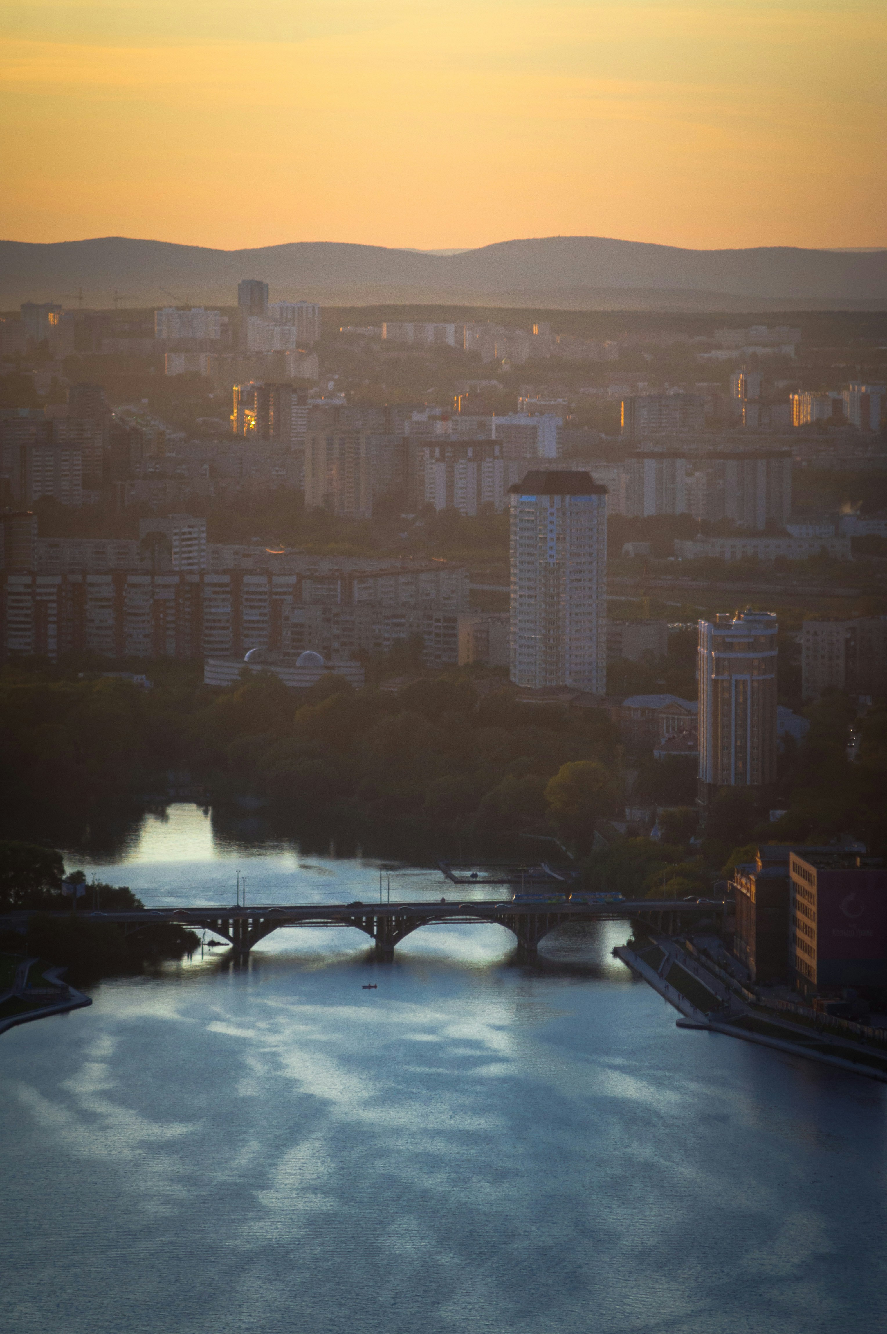 City skyline at sunset with a river reflecting soft golden light and a bridge connecting urban landscapes.