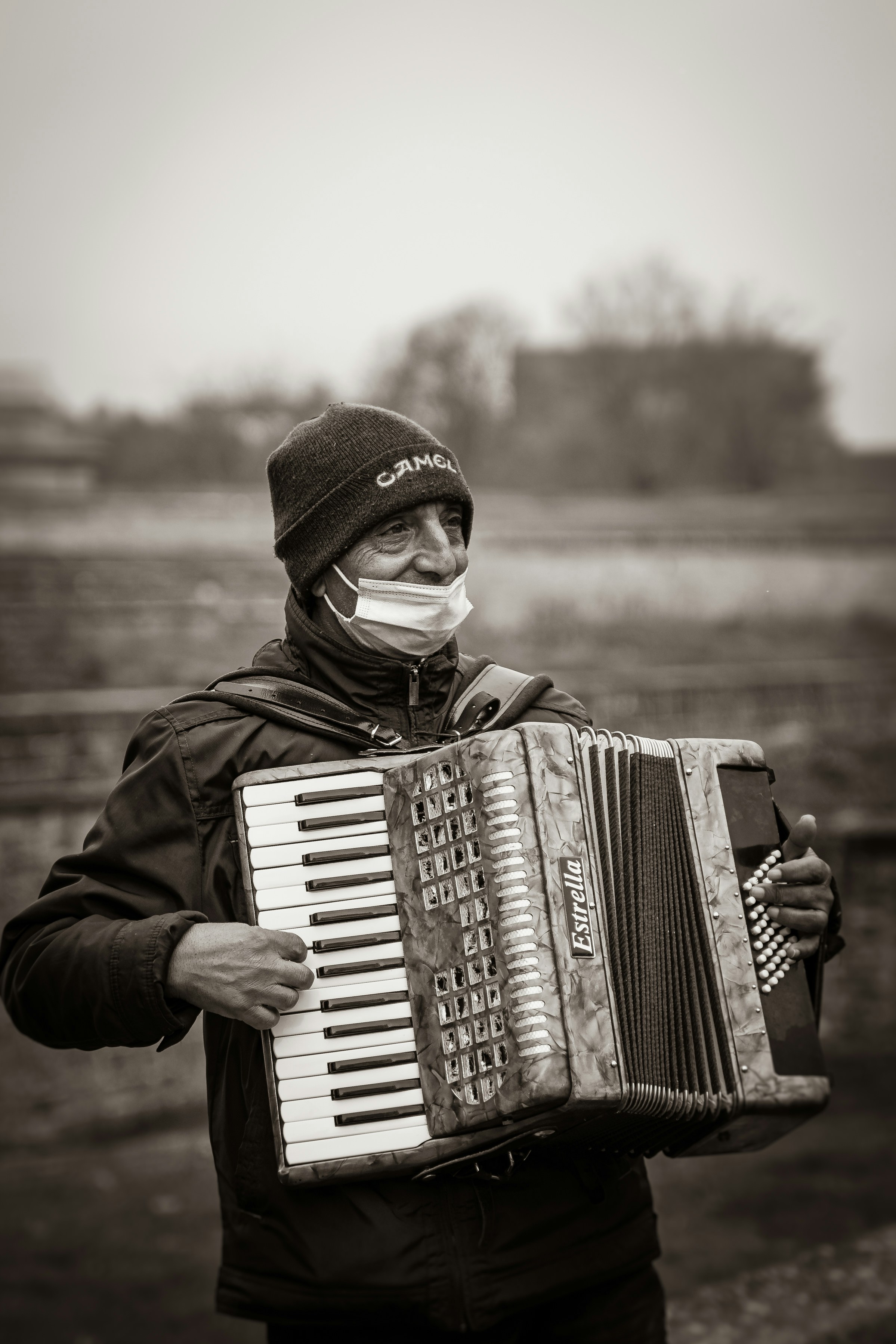 Elderly man playing the accordion in a muted urban setting, wearing a mask and a beanie. His expression reflects a blend of joy and resilience.