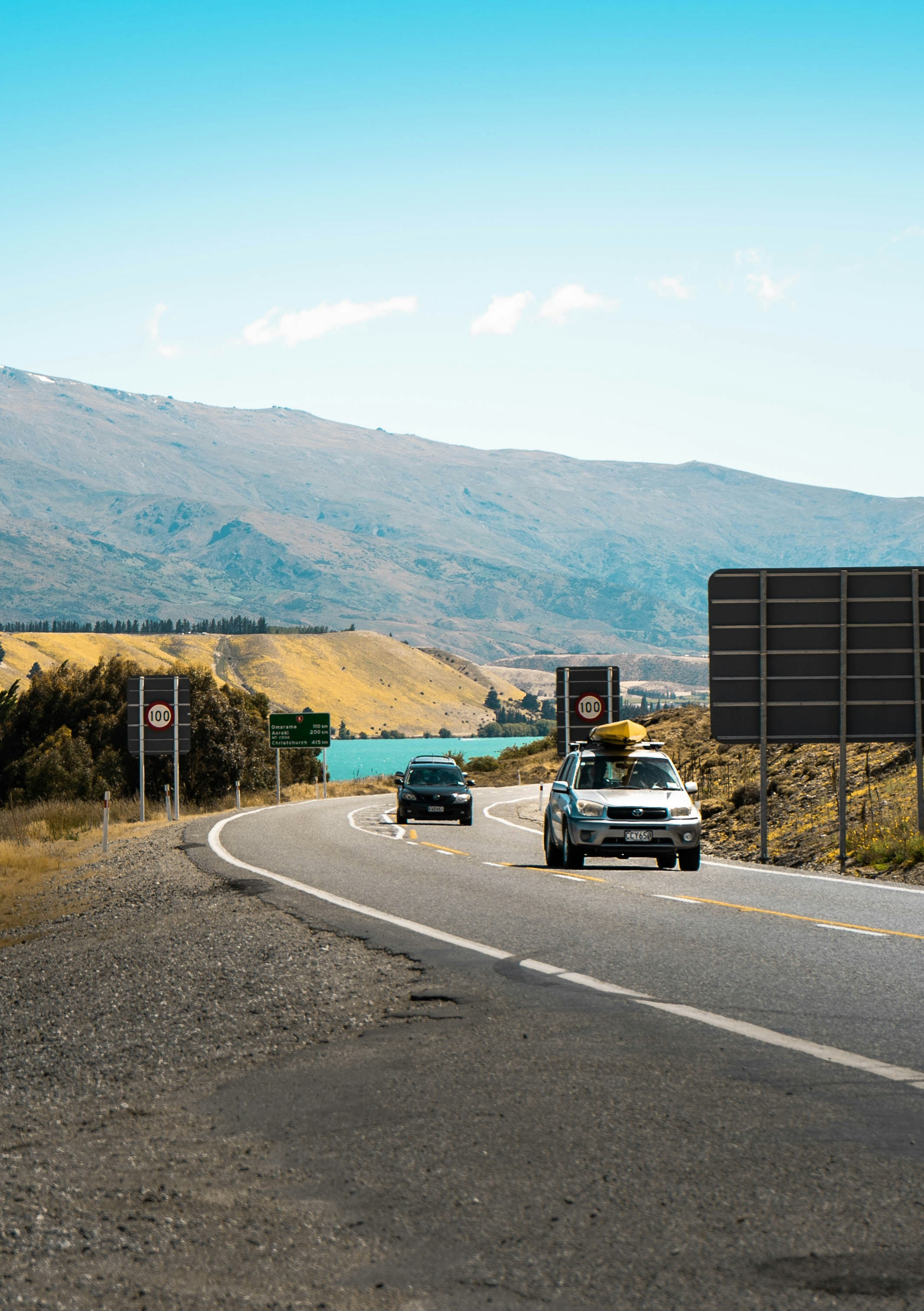 Cars navigate a winding road alongside a tranquil lake, framed by majestic mountains under a clear blue sky.